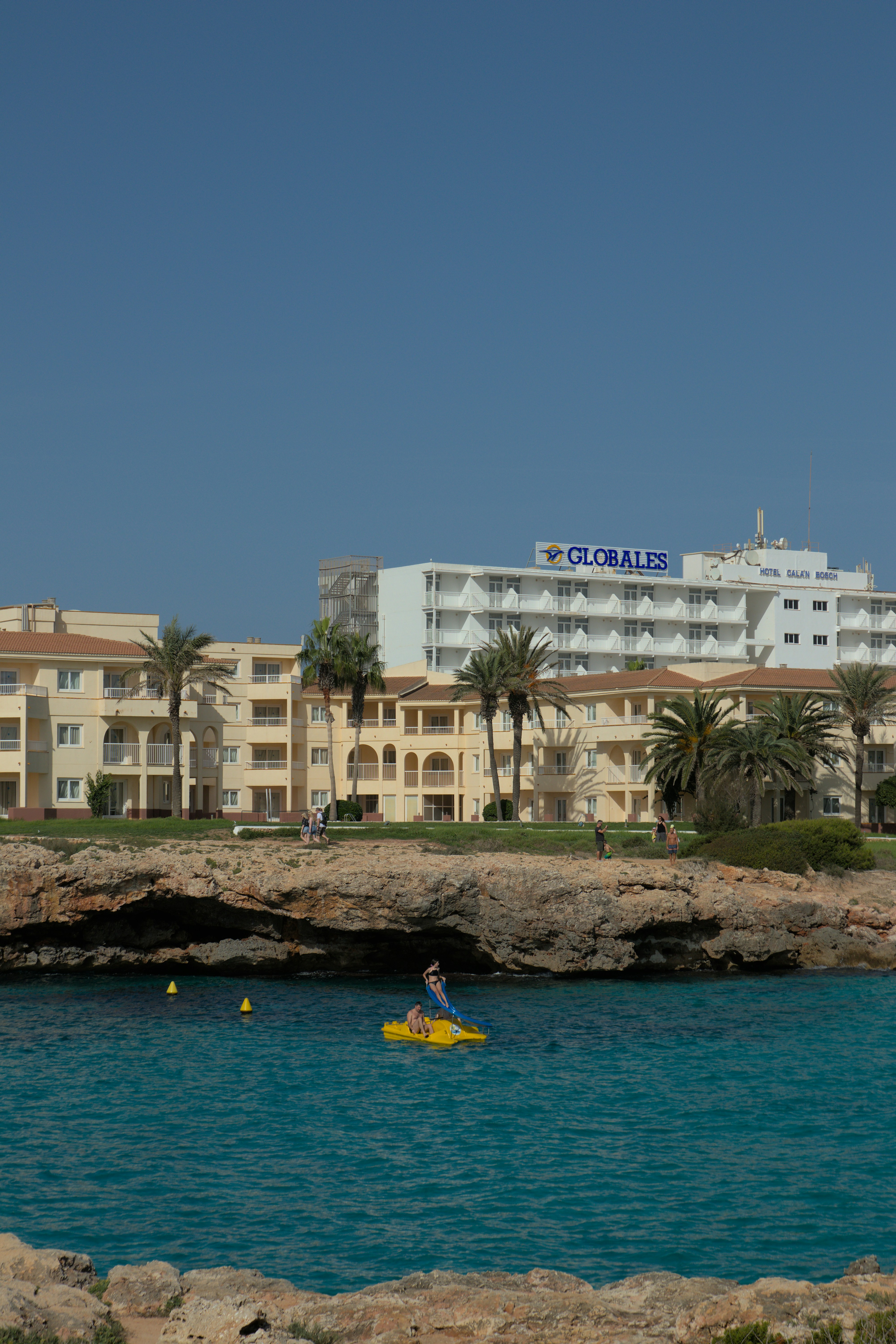 A person enjoys a kayak ride in vibrant blue waters, framed by a coastal resort and palm trees. The scene captures a tranquil day at the beach.