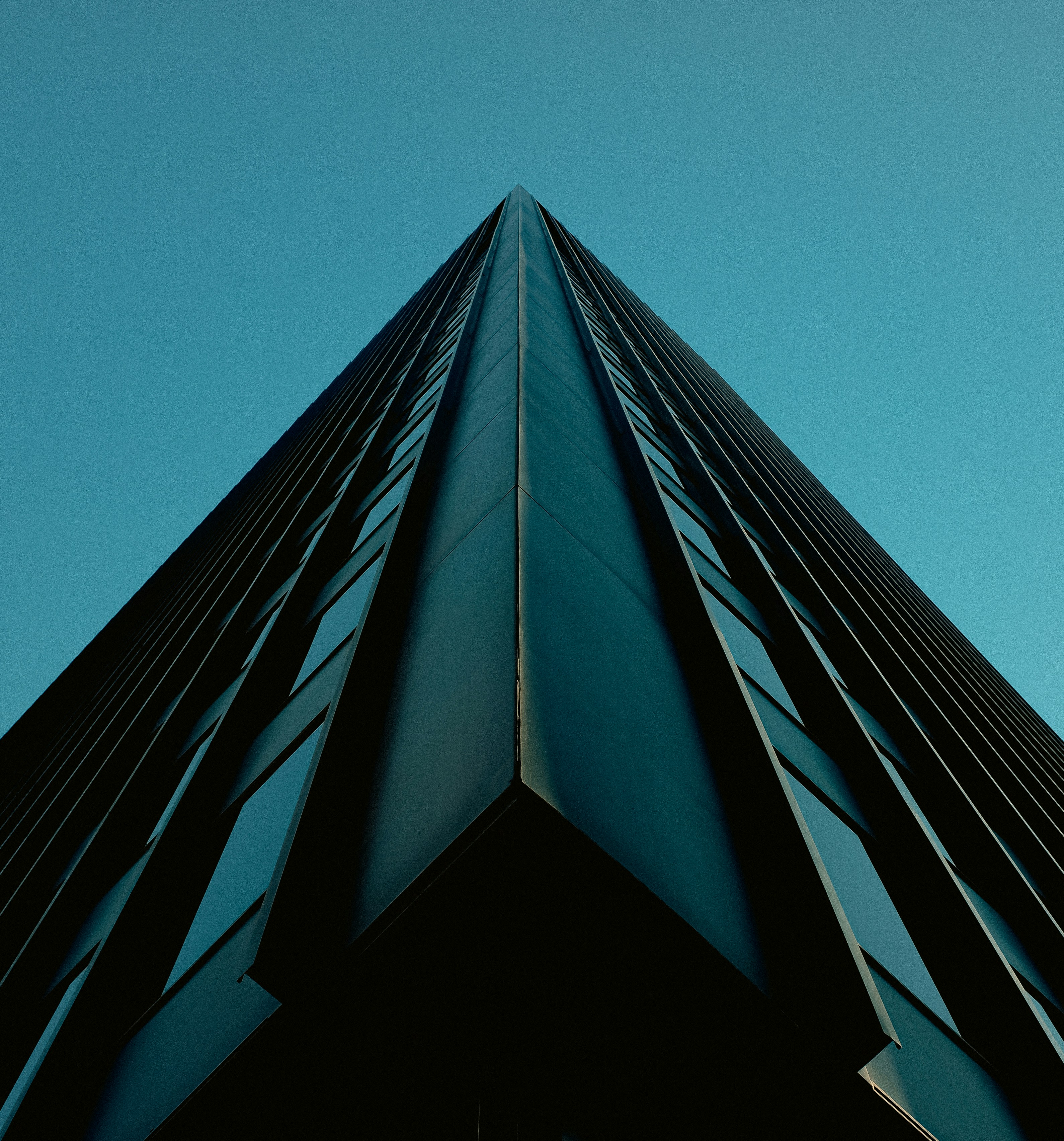 Tall building viewed from the ground, converging into a sharp point against a clear blue sky.