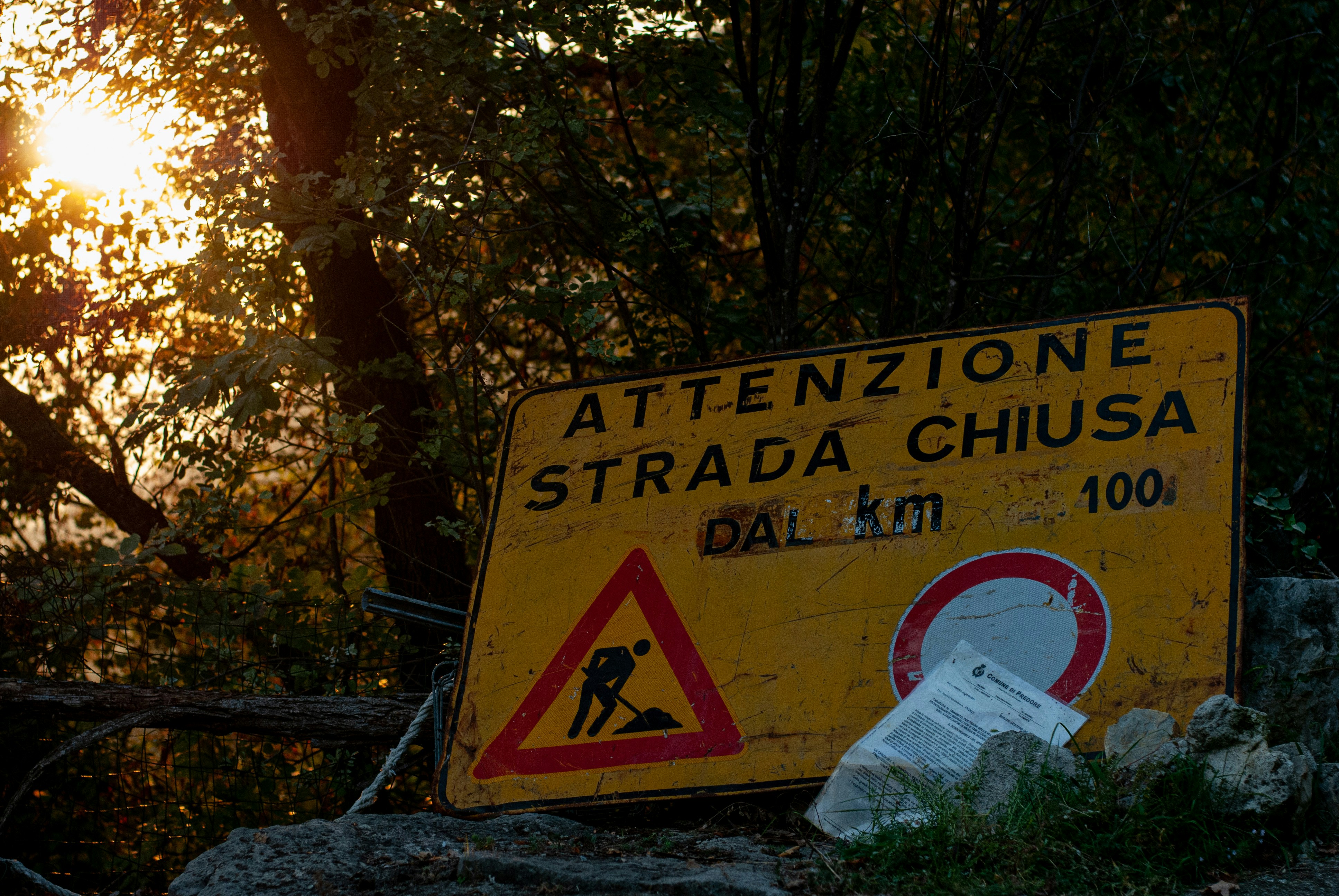 Weathered road sign indicating a closed road, partially obscured by foliage, with a warm sunset glow in the background.