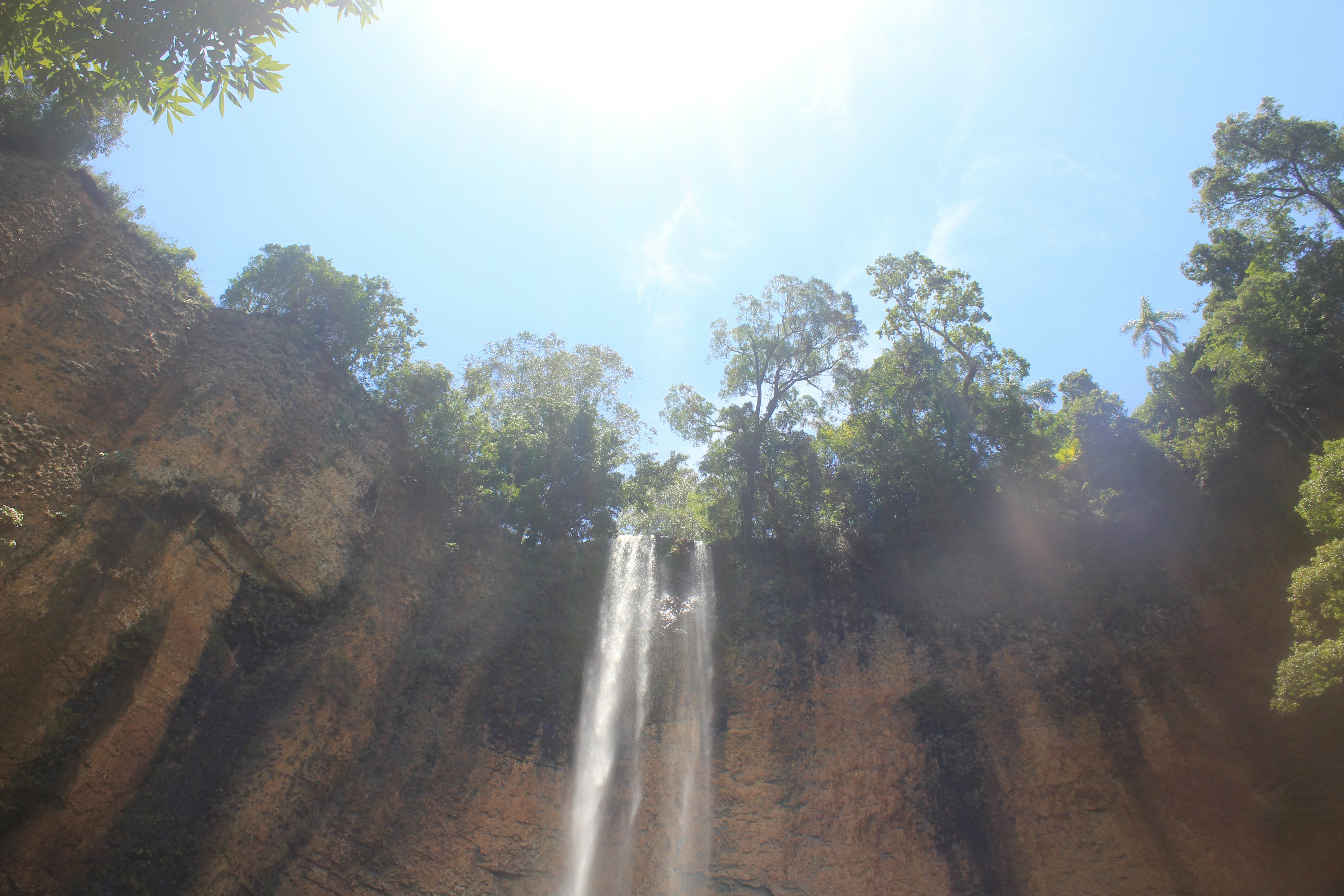 Waterfall cascading down a rocky cliff, framed by lush trees and a bright sky.