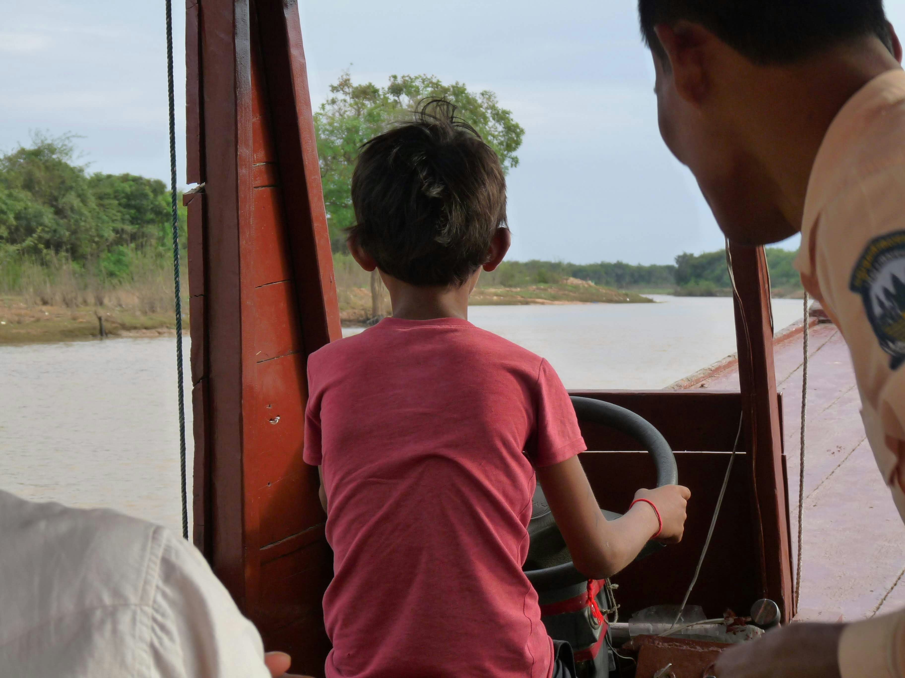 Young boy steering a boat along a tranquil river, flanked by lush greenery and gentle waters.