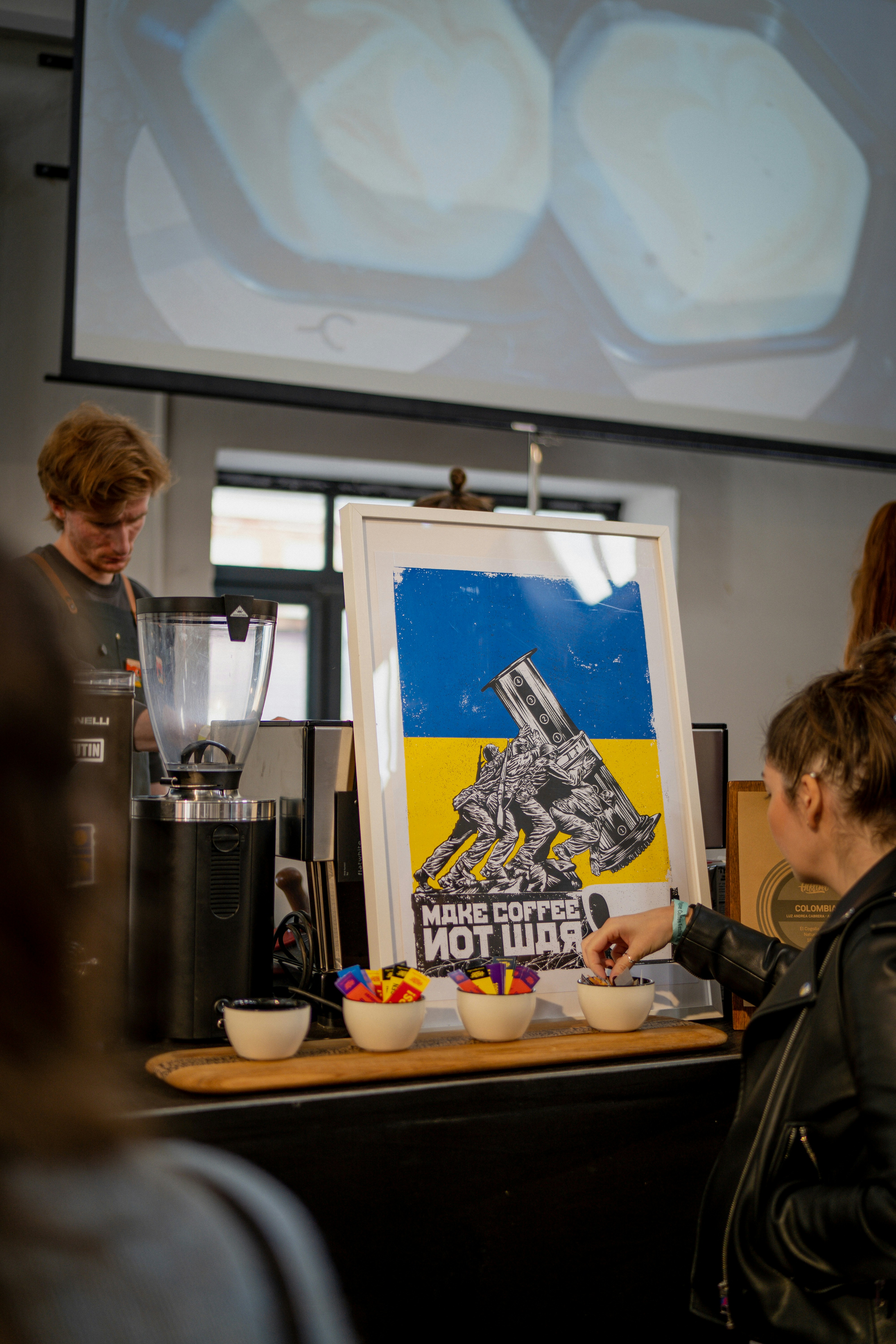Framed illustration on a cafe counter with a blue-yellow flag motif and a stylized scene bearing the slogan MAKE COFFEE NOT WAR.
