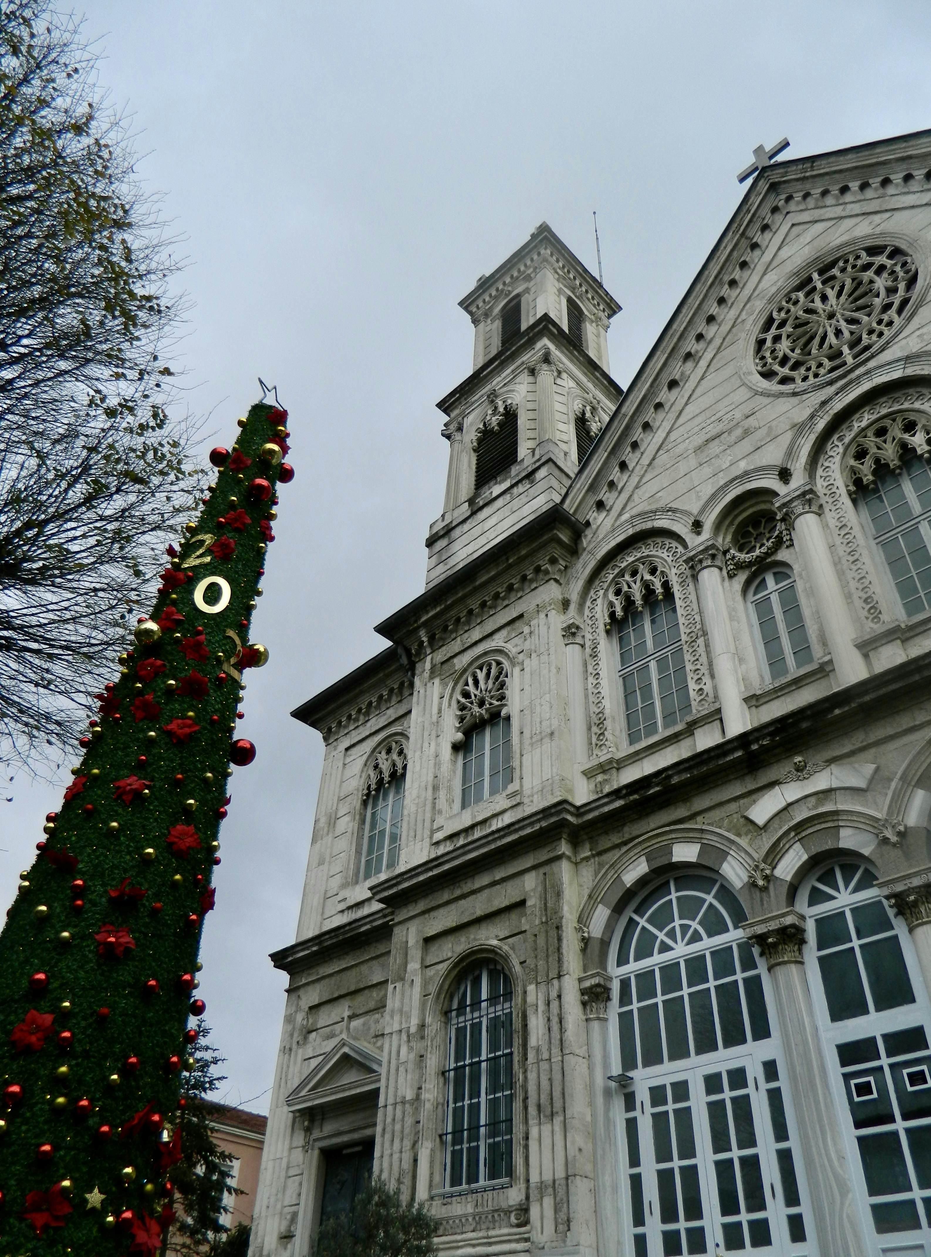 Decorative Christmas tree adorned with ornaments stands beside a historic building featuring intricate architectural details. The scene captures a blend of holiday spirit and cultural heritage.