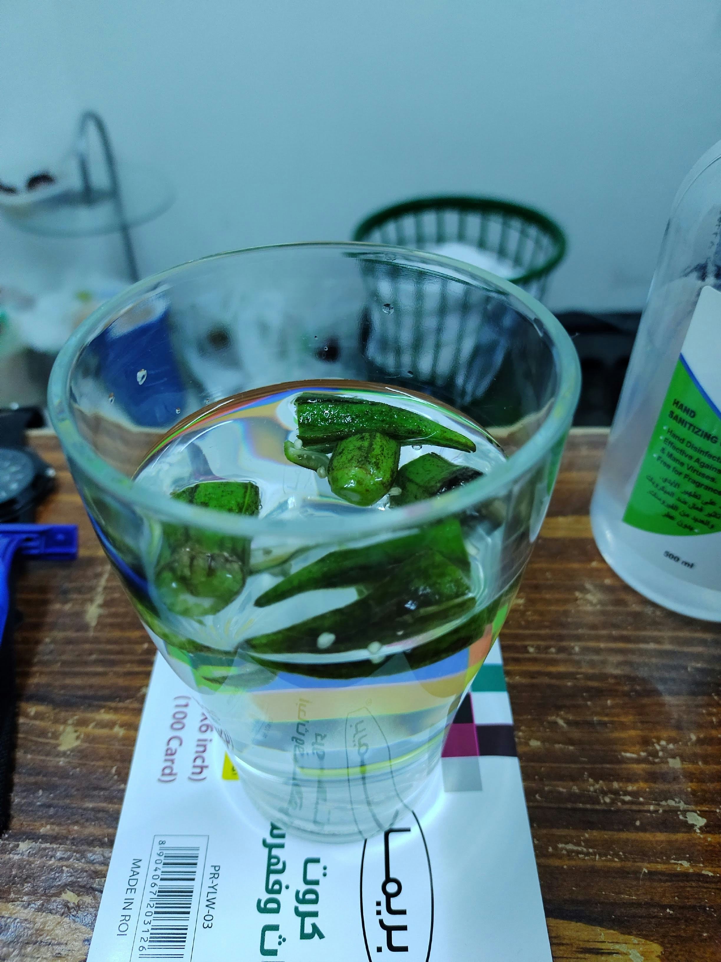 Close-up photograph of a glass with green cucumber pieces floating in water on a wooden counter, with blurred kitchen items in the background.