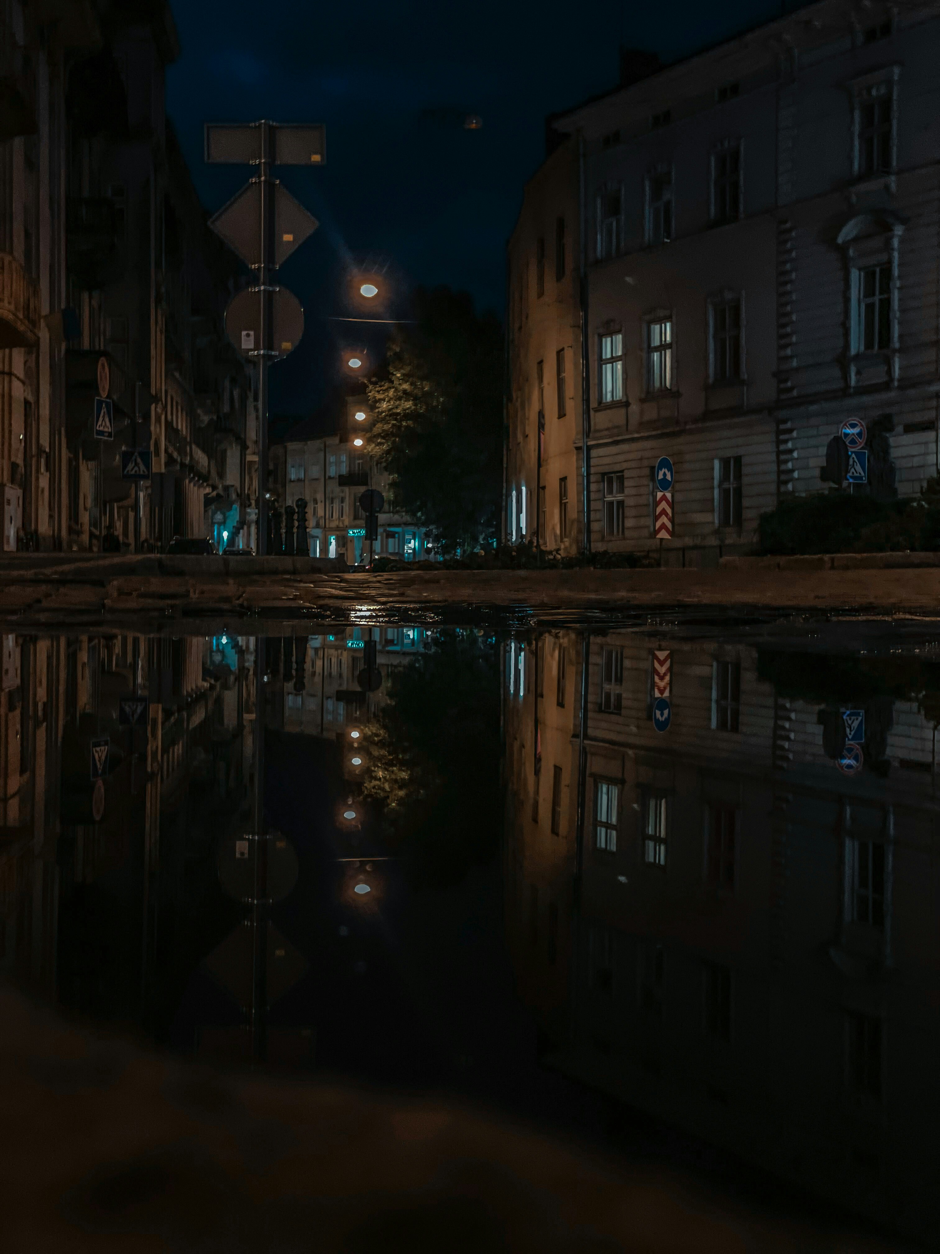 Dimly lit urban street reflecting in a puddle, showcasing the interplay of light and architecture at night.