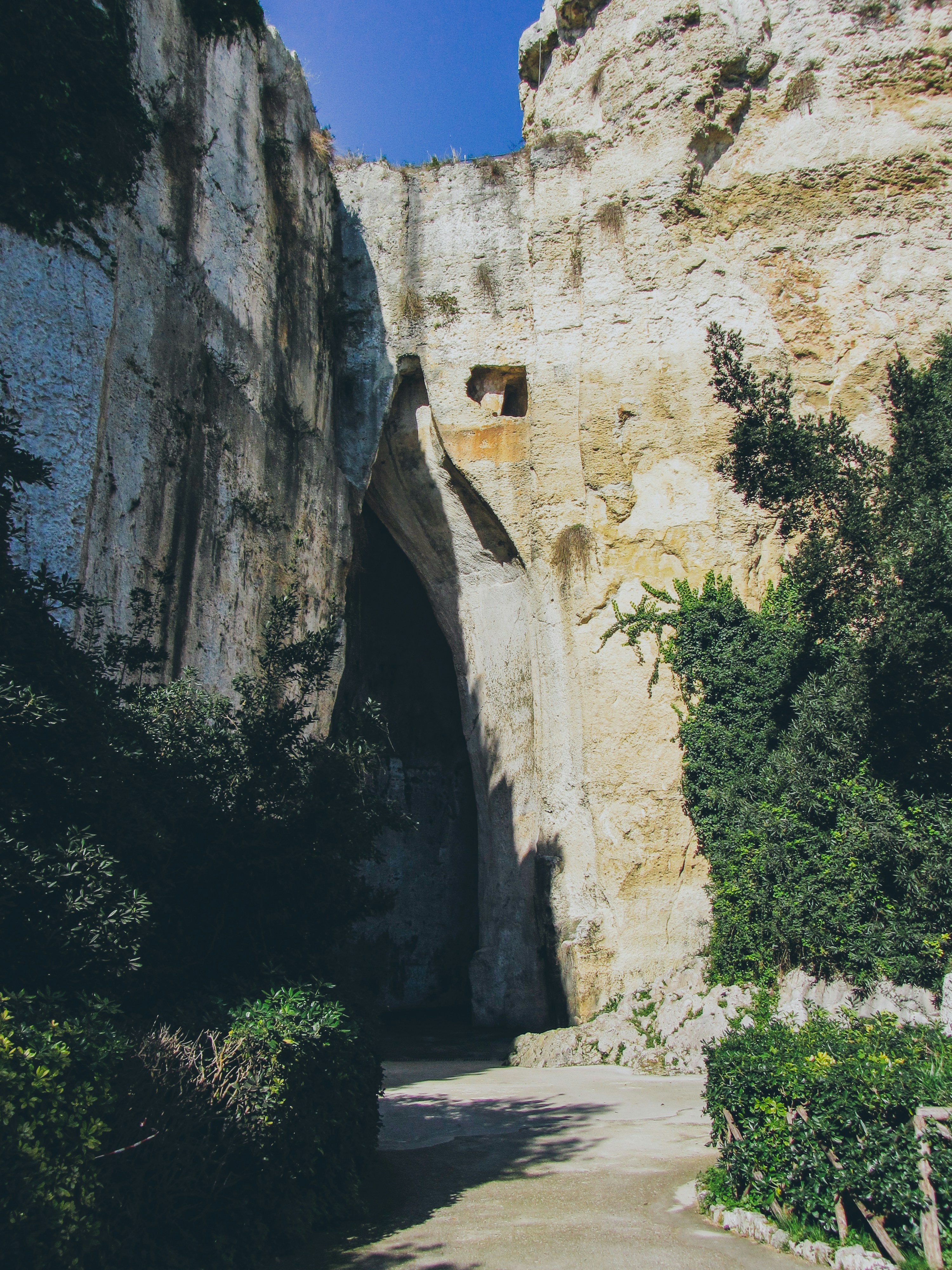A sunlit cliff arch frames a dark cave entrance along a sandy path. Lush shrubs cling to the rock walls, highlighting the contrast between light and shadow.