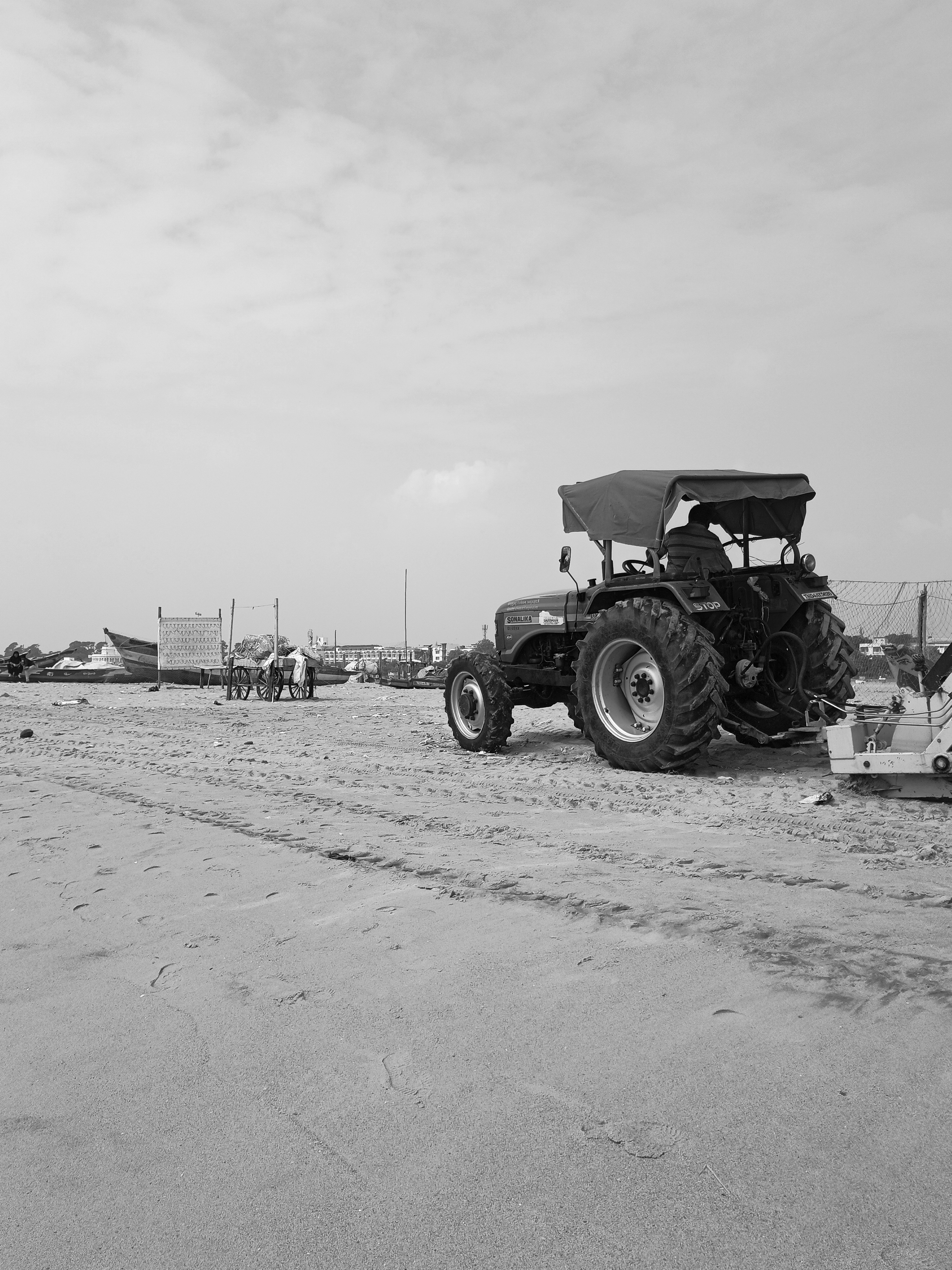 A vintage tractor parked on a sandy beach, with fishing boats and people in the background. The scene captures the essence of coastal life.