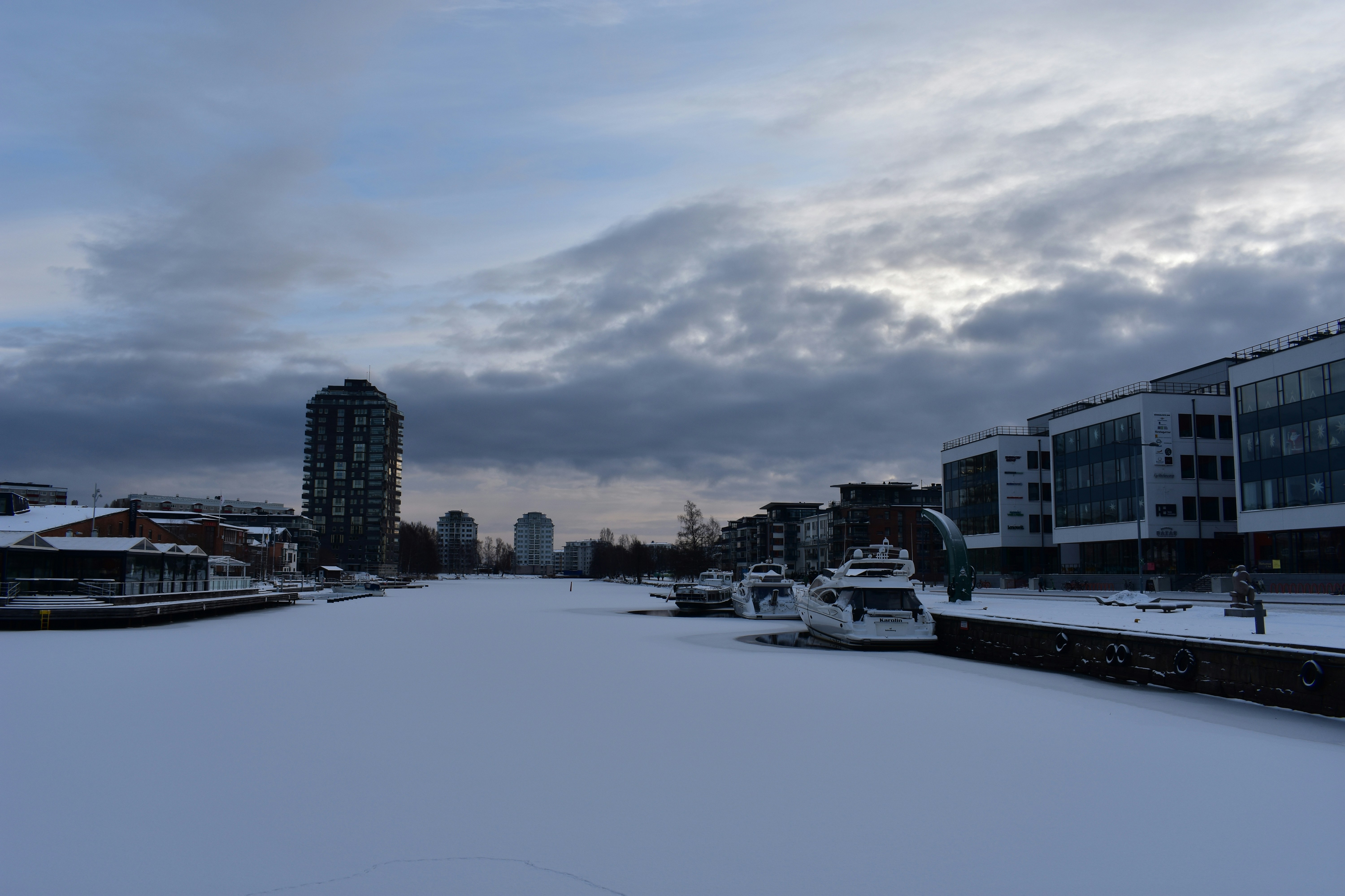 Snow-covered urban riverfront with modern buildings and boats under a cloudy winter sky.