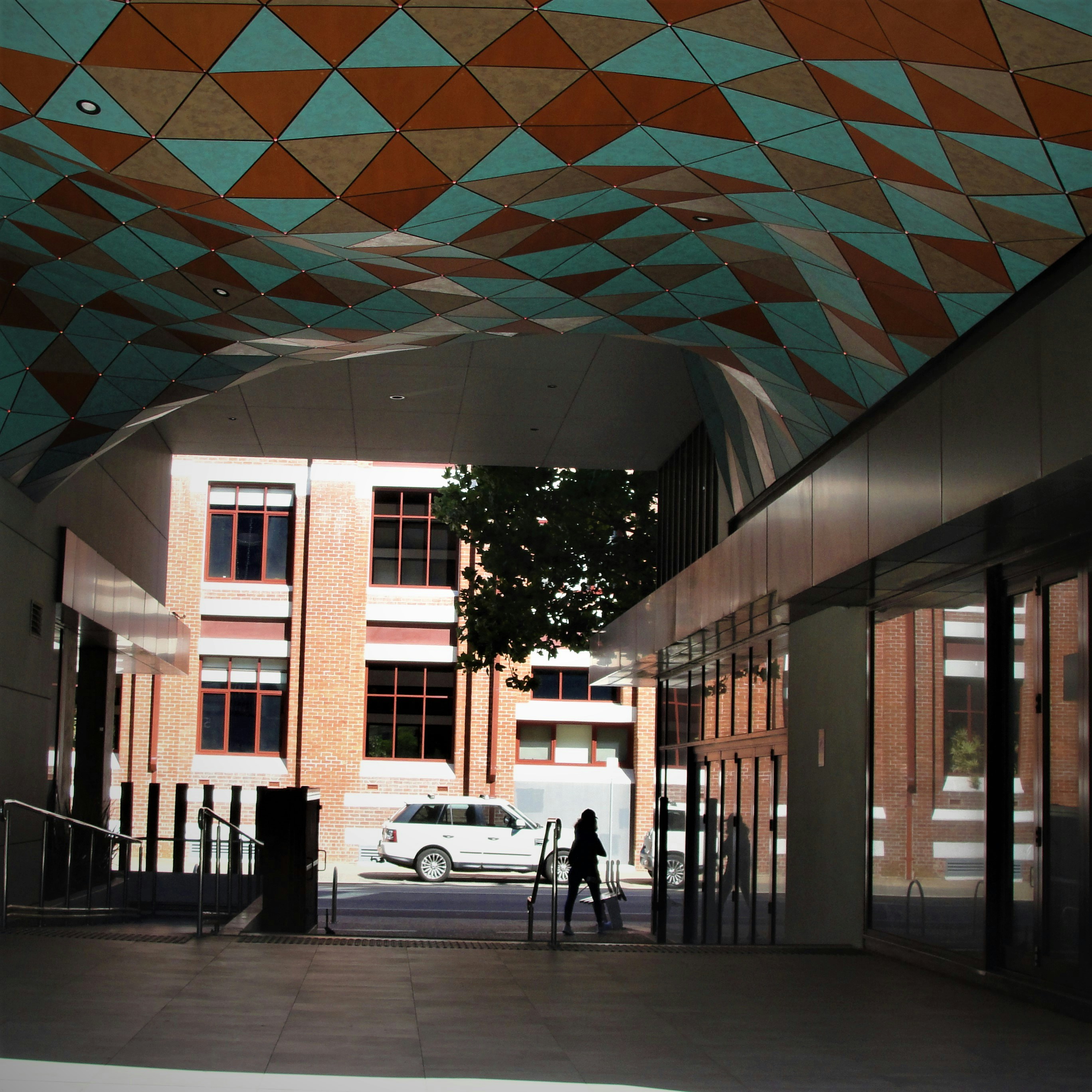 Under a tessellated teal and orange canopy, a brick courtyard opens to a sunlit street where two silhouettes walk past a parked car.
