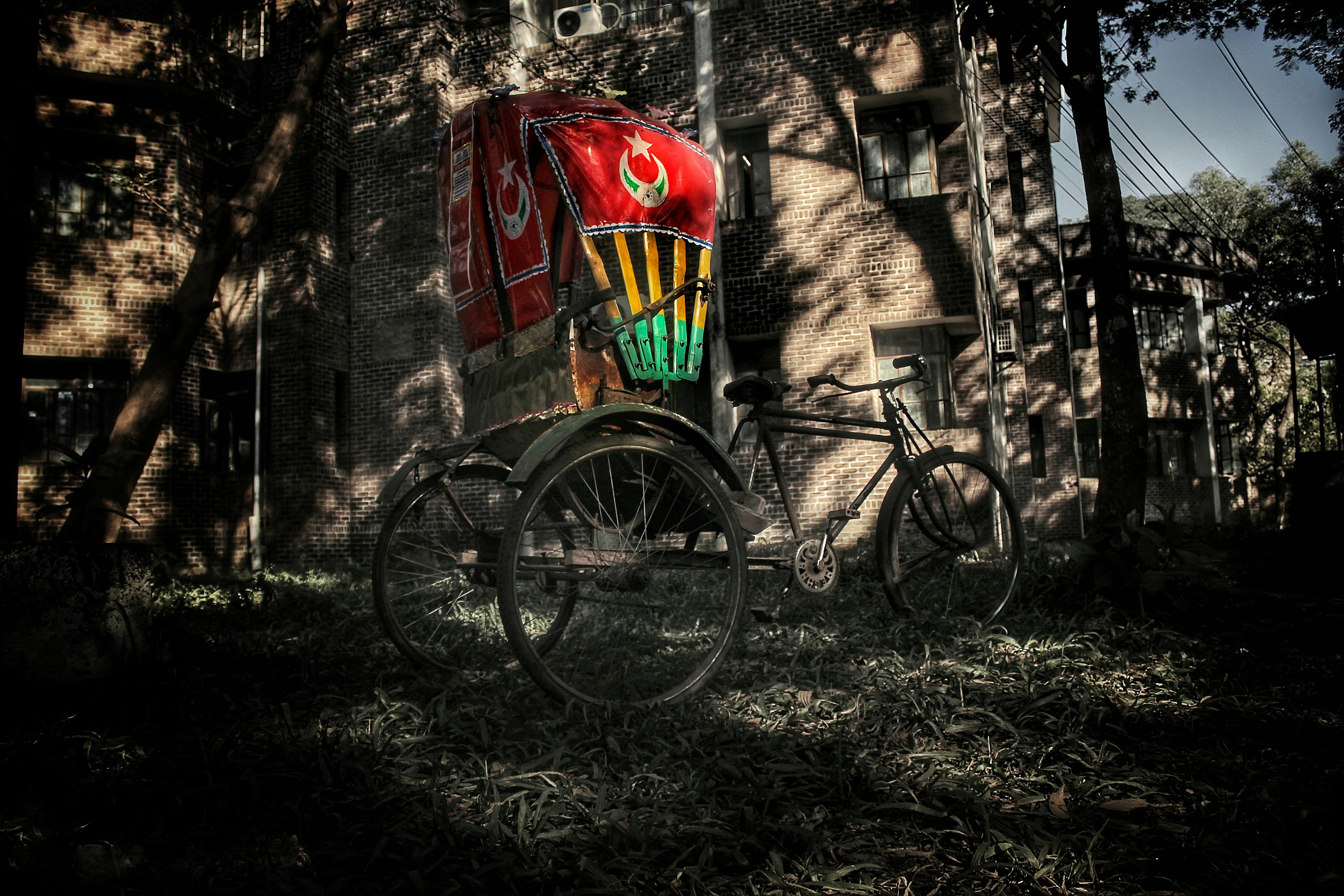 Photograph of a bicycle rickshaw with a colorful canopy parked in a shadowy, sun-dappled urban yard.