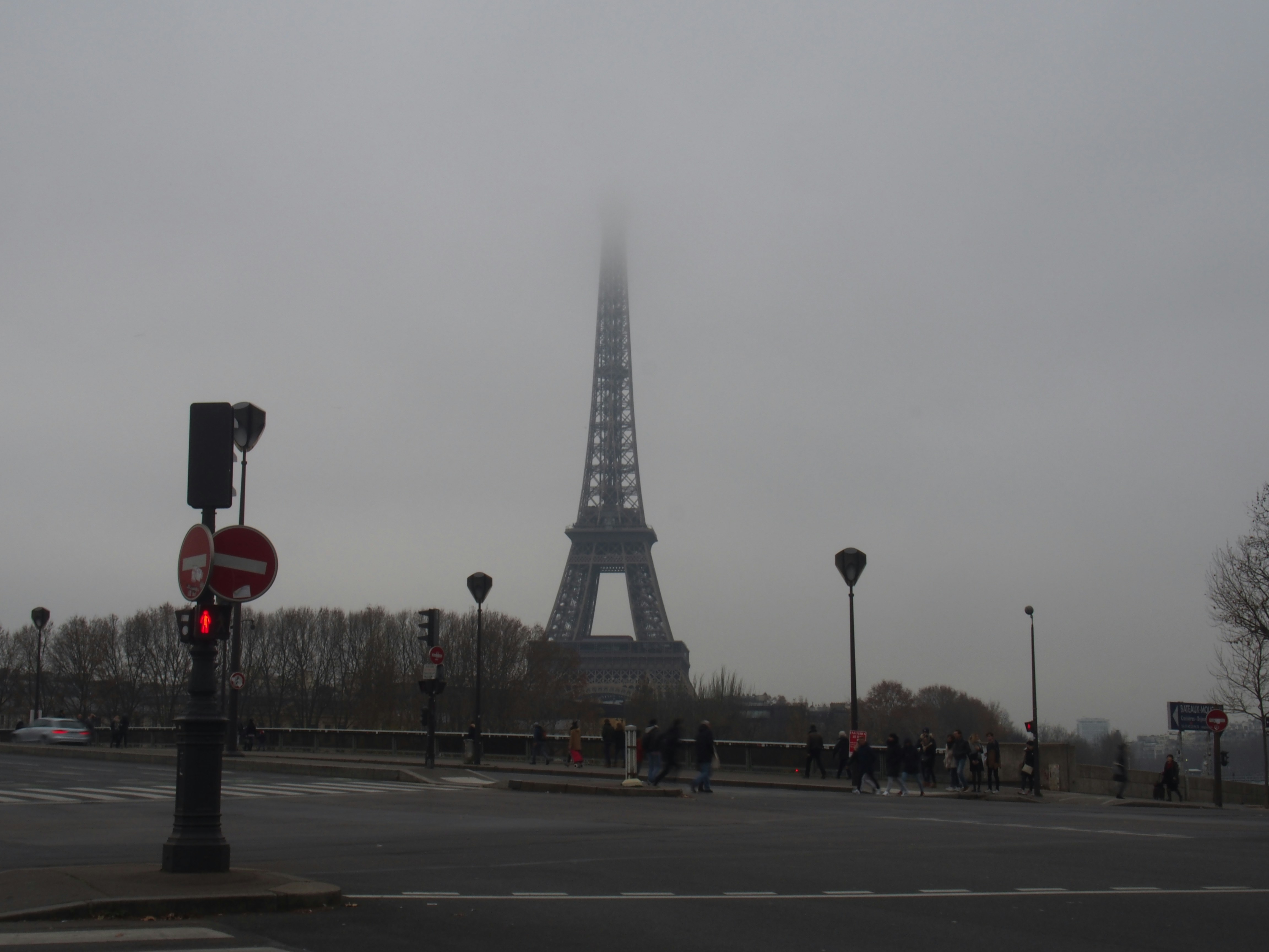 Eiffel Tower partially obscured by fog, creating a mysterious atmosphere. Pedestrians and traffic signals add context to the scene.
