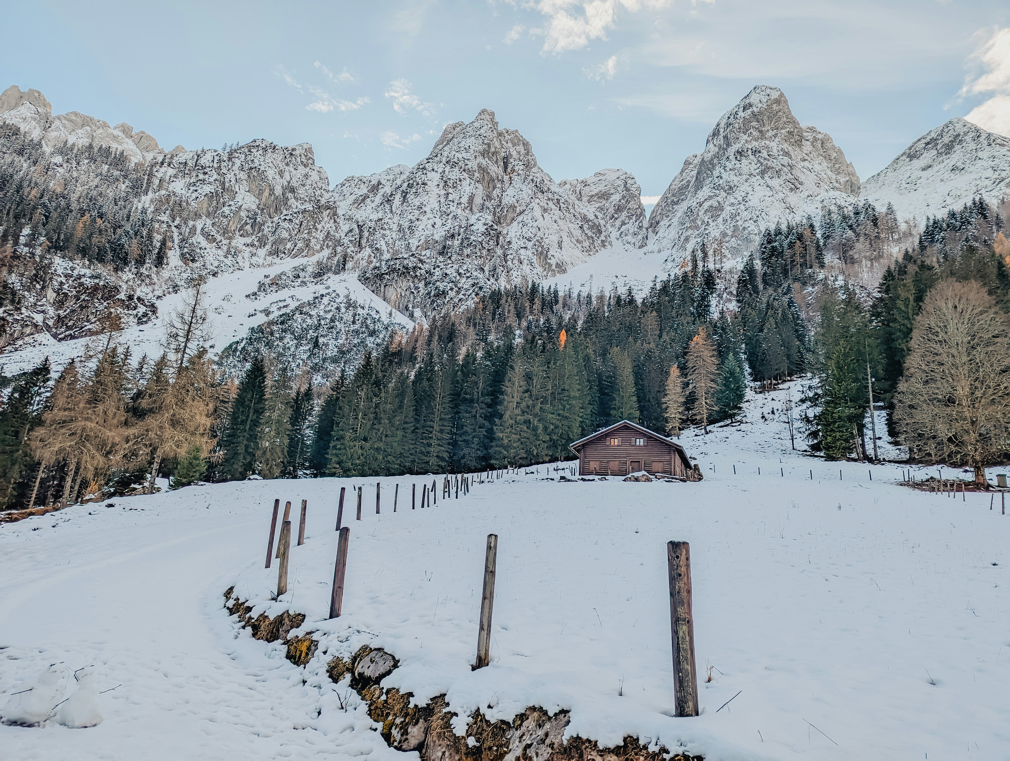 Snow-covered alpine valley with a wooden chalet, fence posts, evergreens, and towering snow-capped mountains in the background.