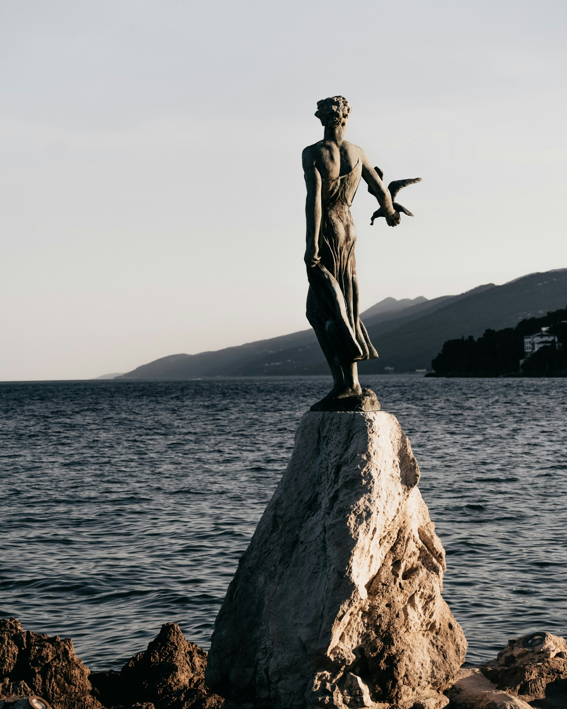 A solitary bronze statue stands atop a rocky outcrop, gazing over a tranquil sea under a soft sky. The scene captures the serene connection between art and nature.