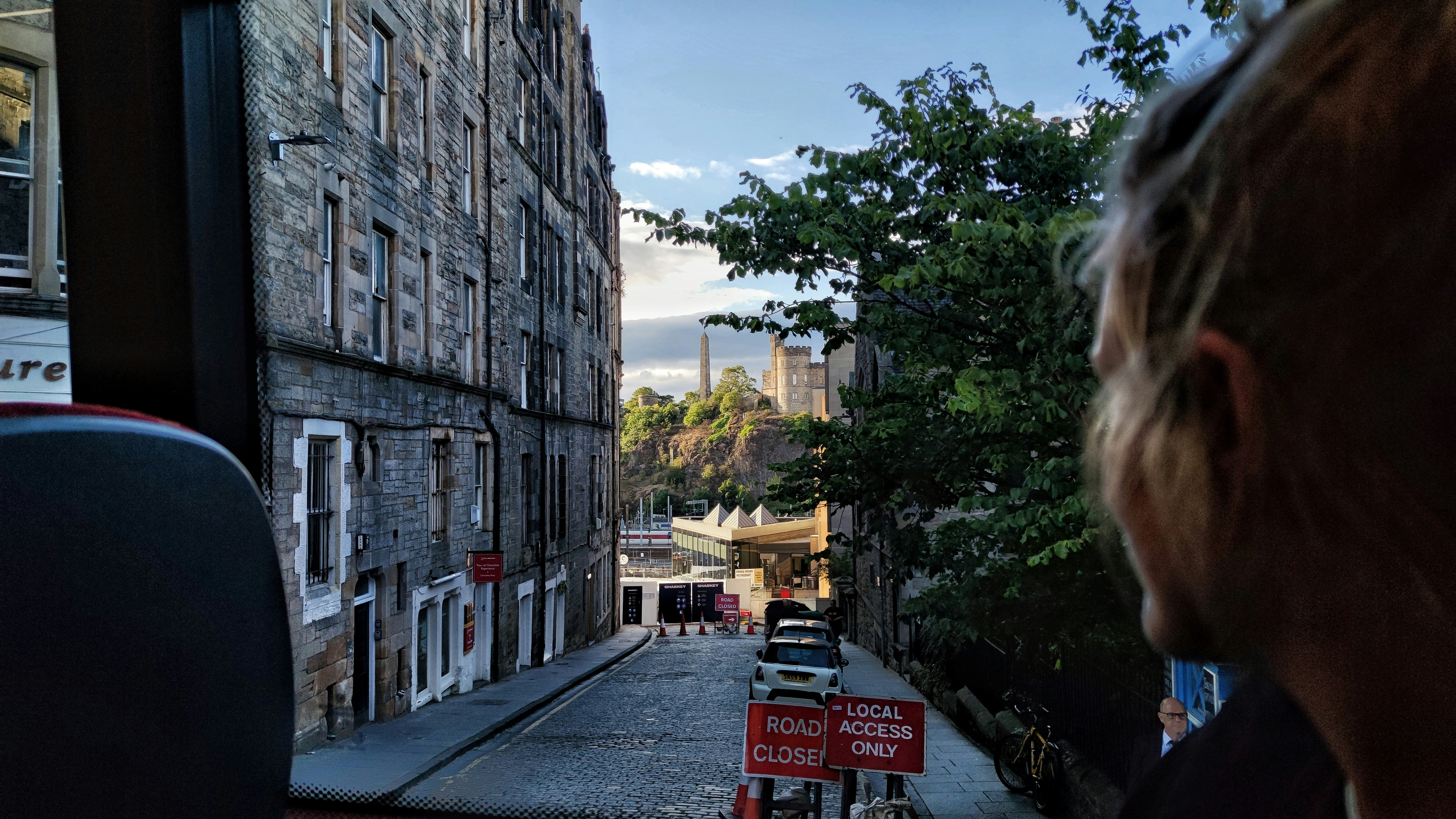 A narrow street lined with stone buildings leads toward a distant town center, framed by a tree on the right and a foreground figure.