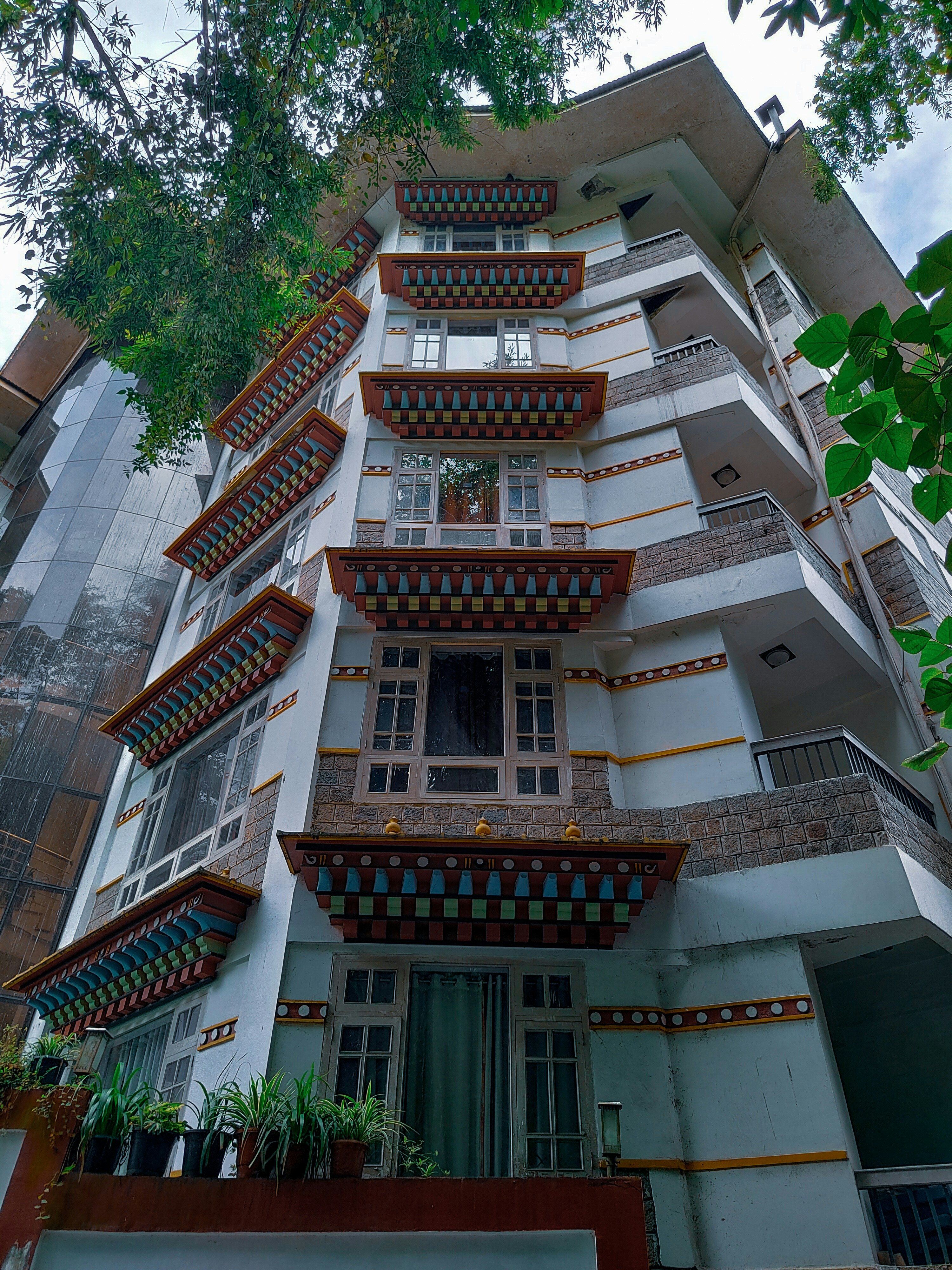 A multi-story building with stacked, pagoda-inspired balconies painted in vibrant colors, forming a tiered facade. Greenery frames the lower level, while the angular rooflines create a distinctive skyline.