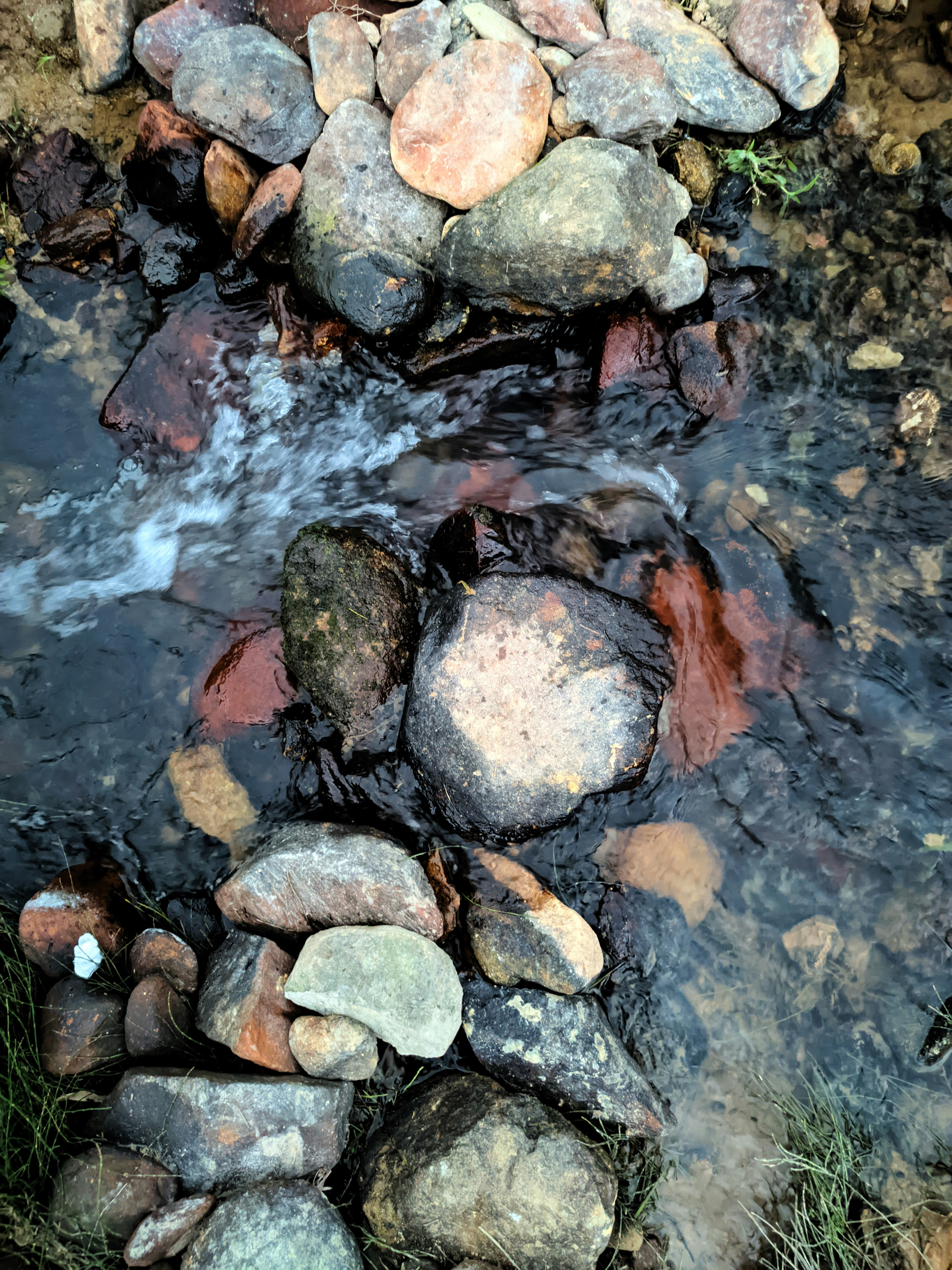 A serene stream flowing over a bed of colorful stones, showcasing the intricate patterns formed by nature. The water glistens as it dances around the rocks.