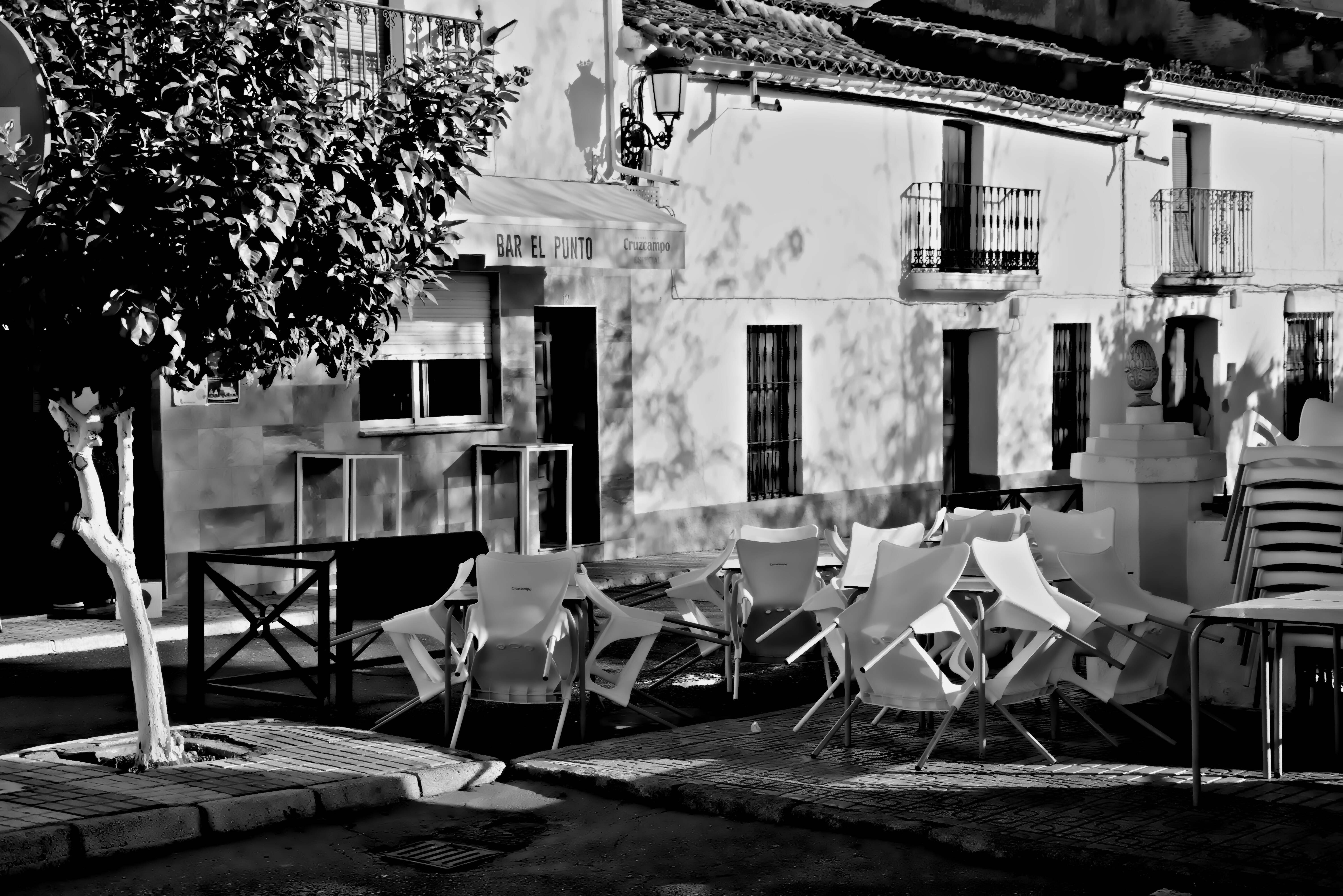 Black and white scene of an empty outdoor café with chairs and tables under a tree's shadow in a small village.