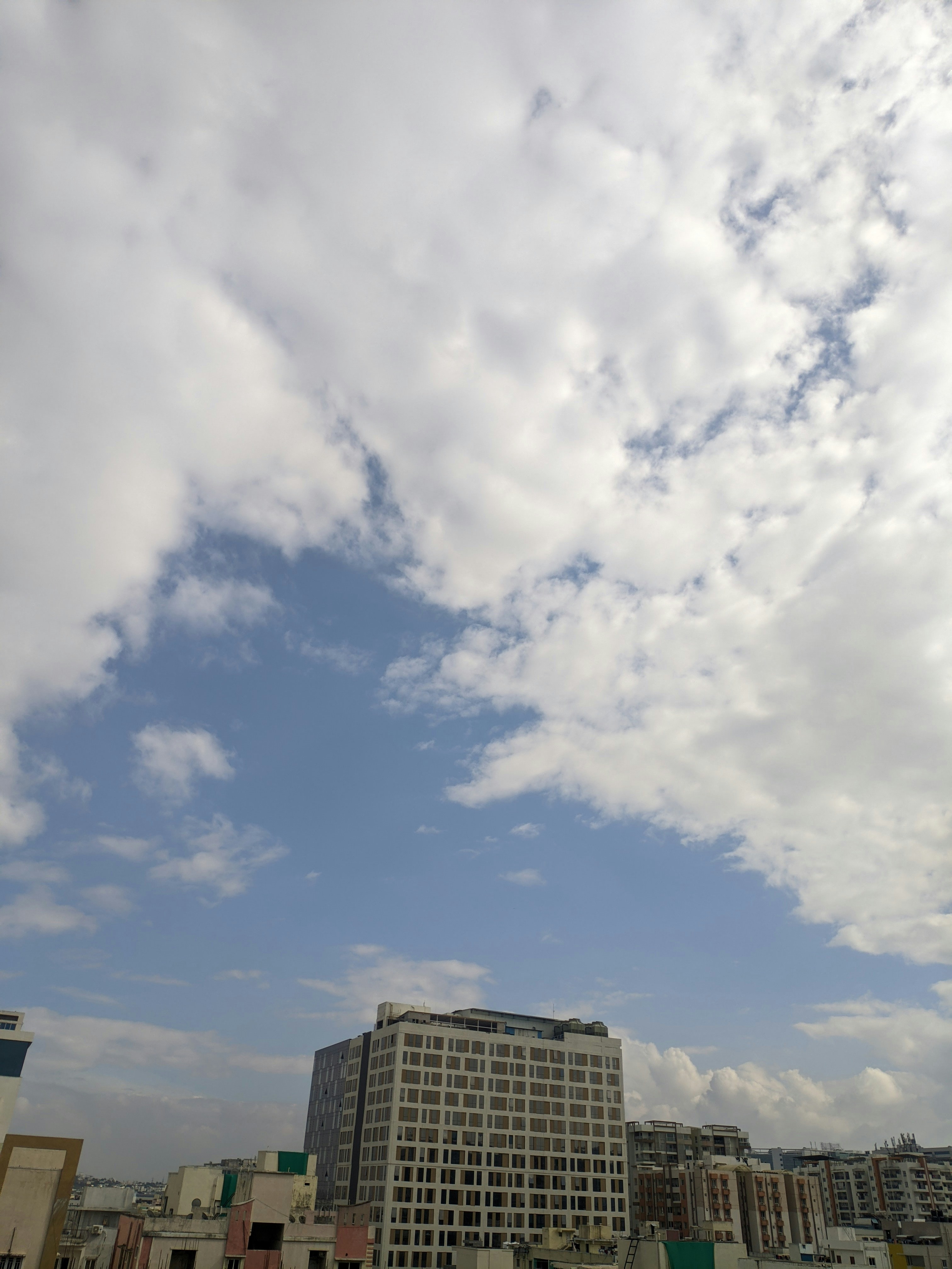 Low-angle view of a city skyline with a bright, cloud-filled sky above; a large modern building anchors the foreground, with smaller structures lining the base.
