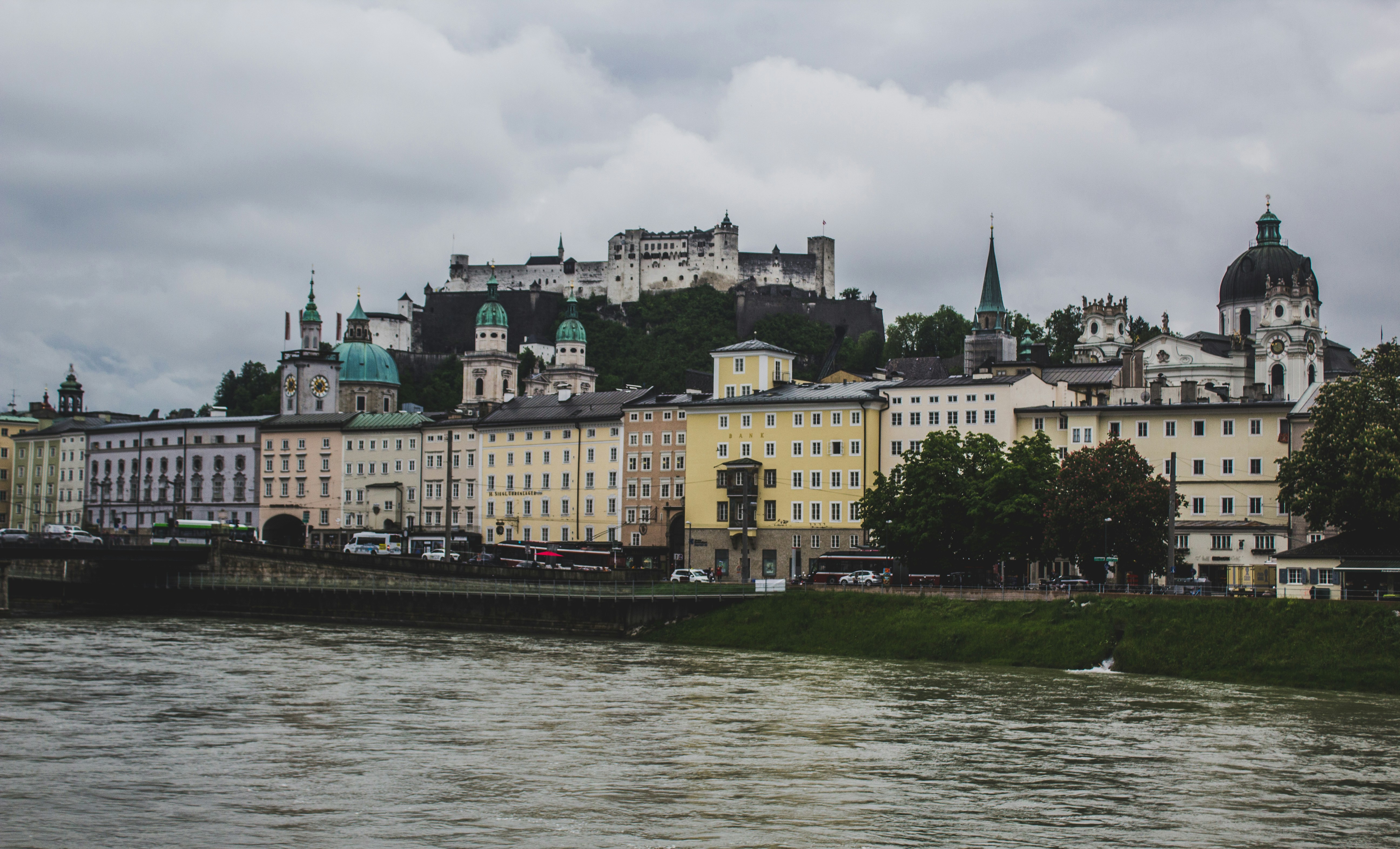 Charming riverside architecture of Salzburg under a moody sky, with the Hohensalzburg Fortress perched above. The scene captures the city's rich history and vibrant colors.