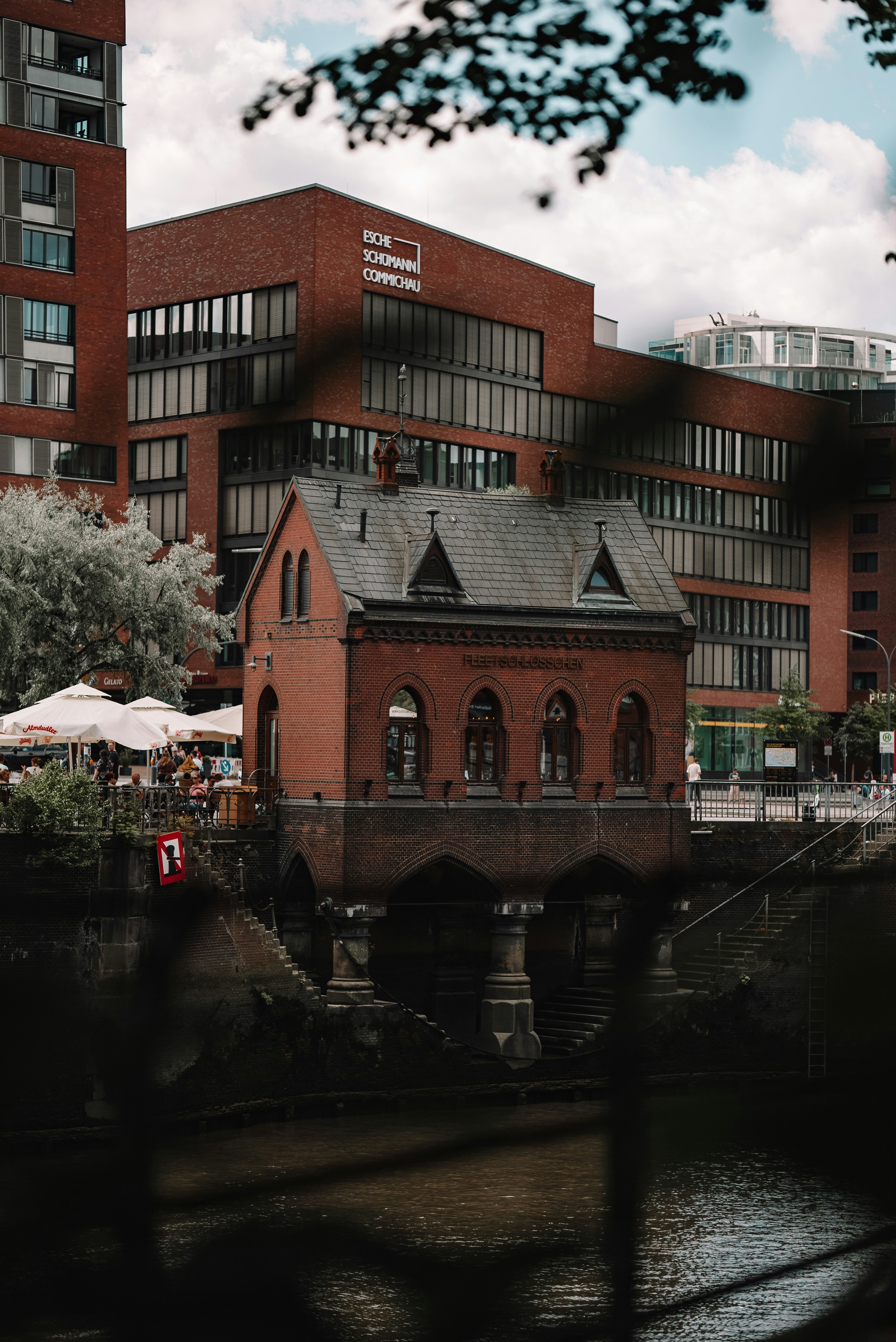 Historic brick riverside house with arched windows sits along a canal, framed by a modern urban backdrop of brick and glass buildings. Foreground railing silhouettes frame the lower edge, adding depth.