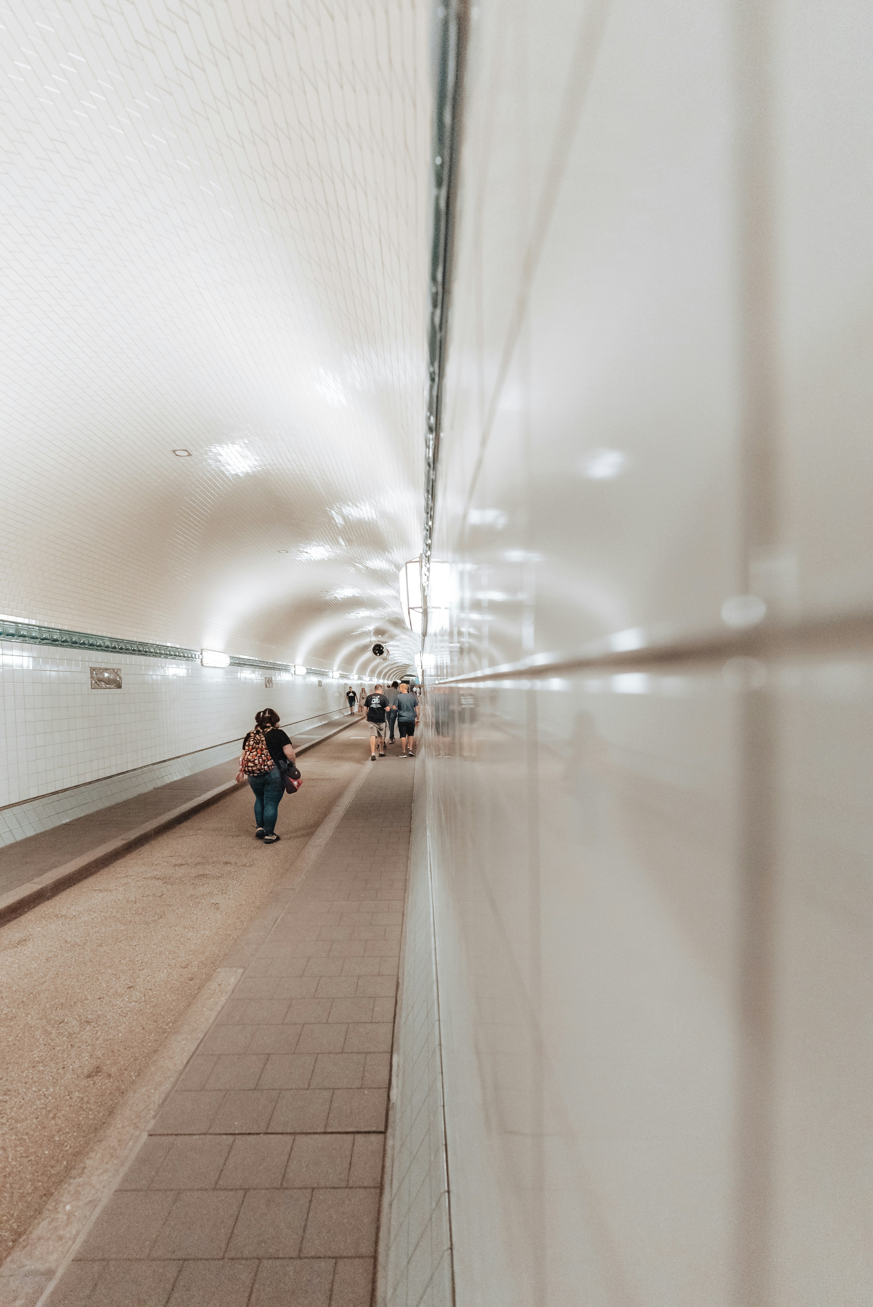 Long, white-tiled tunnel with a central walkway and converging lines toward a bright vanishing point. A small group of pedestrians walks away from the camera, adding scale and depth.