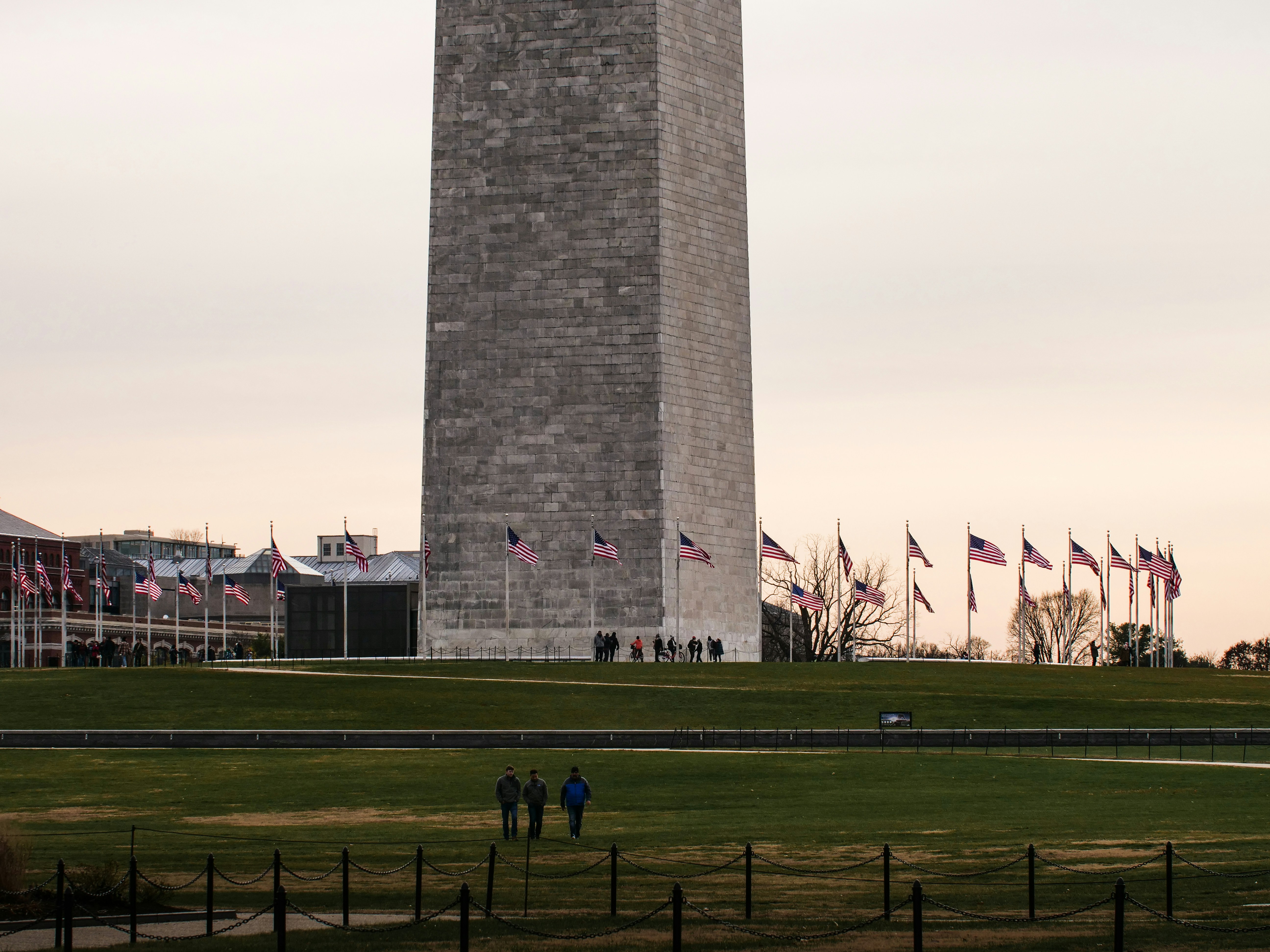 Washington Monument stands tall against a twilight sky, flanked by waving flags and a few silhouetted visitors. The scene evokes a sense of history and reflection.