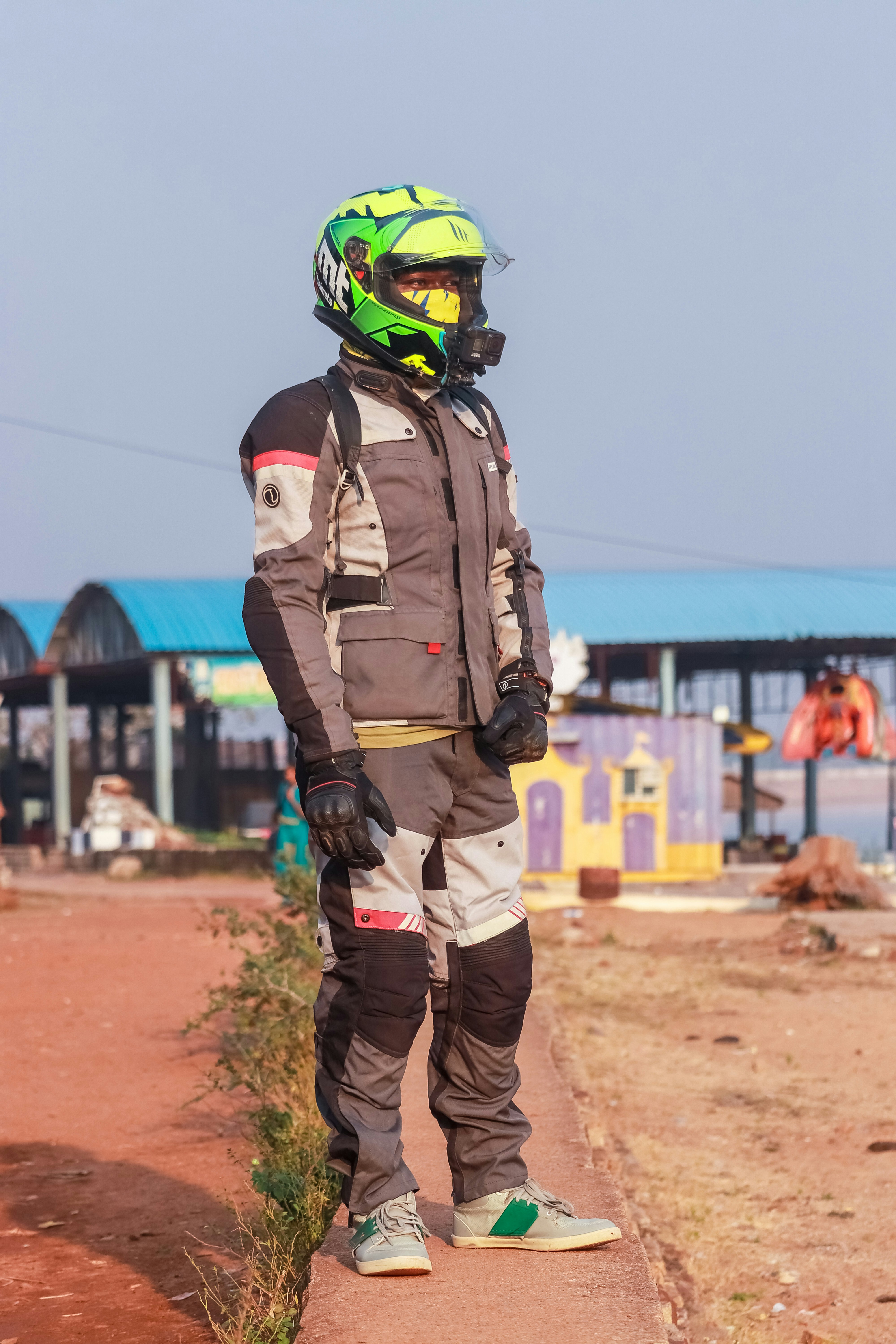 Motorcyclist in full gear stands on a concrete ledge with buildings in the background under a clear sky.
