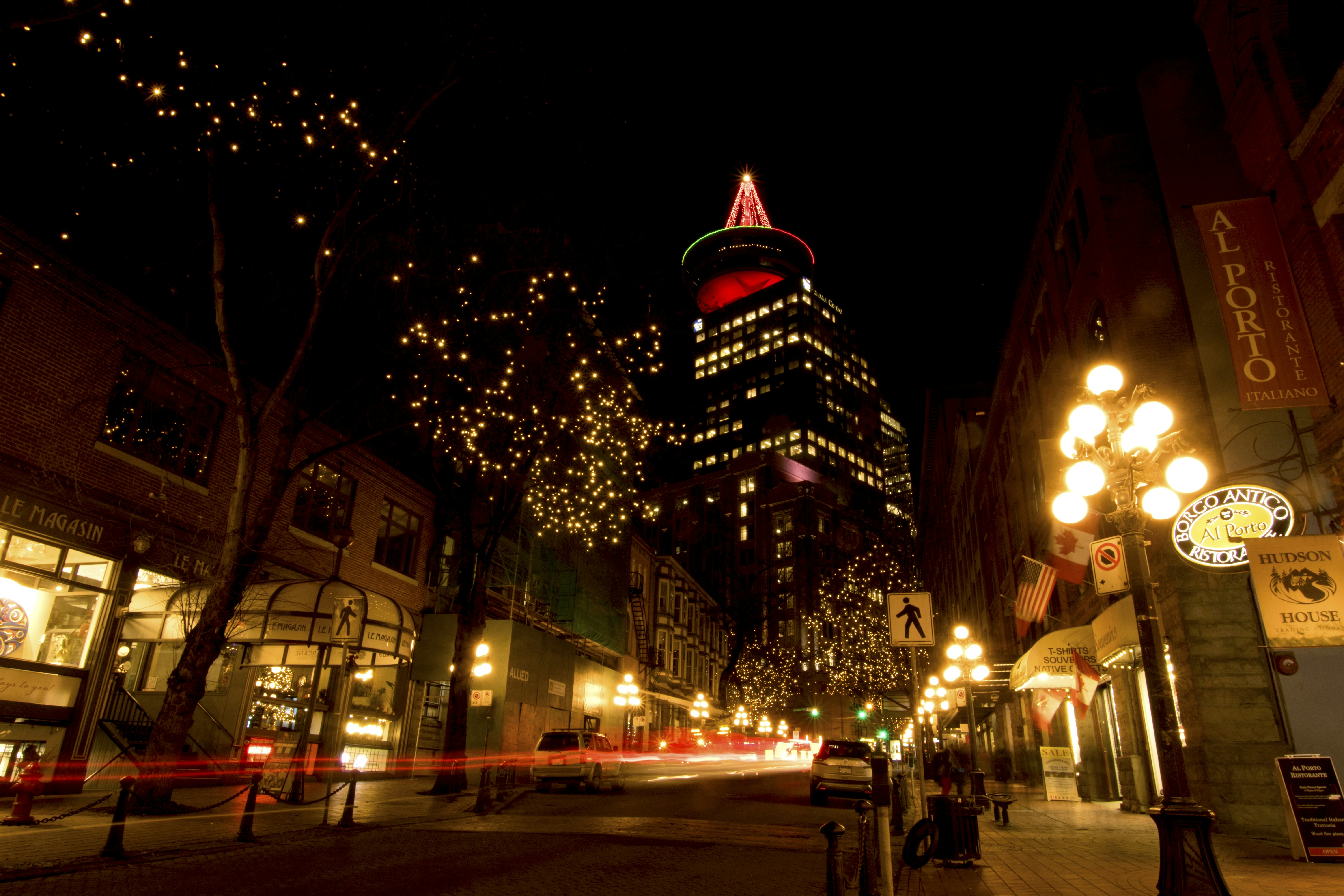 Night street scene with string lights overhead, a tall central building with a red-lit spire, and light trails from passing cars along a storefront-lined boulevard.