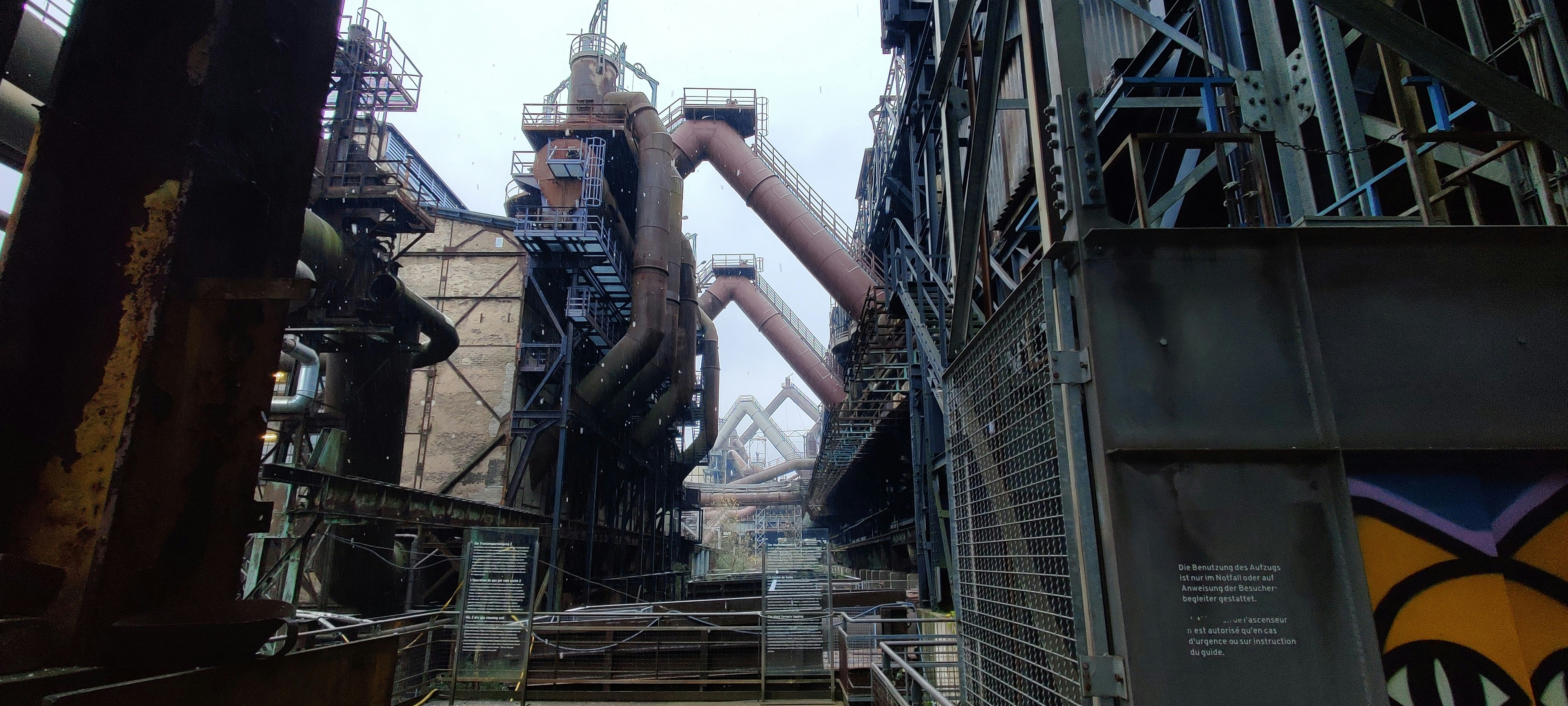 Industrial facility with pipes and steel structures converging in the frame; foreground shows weathered metalwork and a hint of colorful wall art, capturing an expansive, gritty workplace scene.