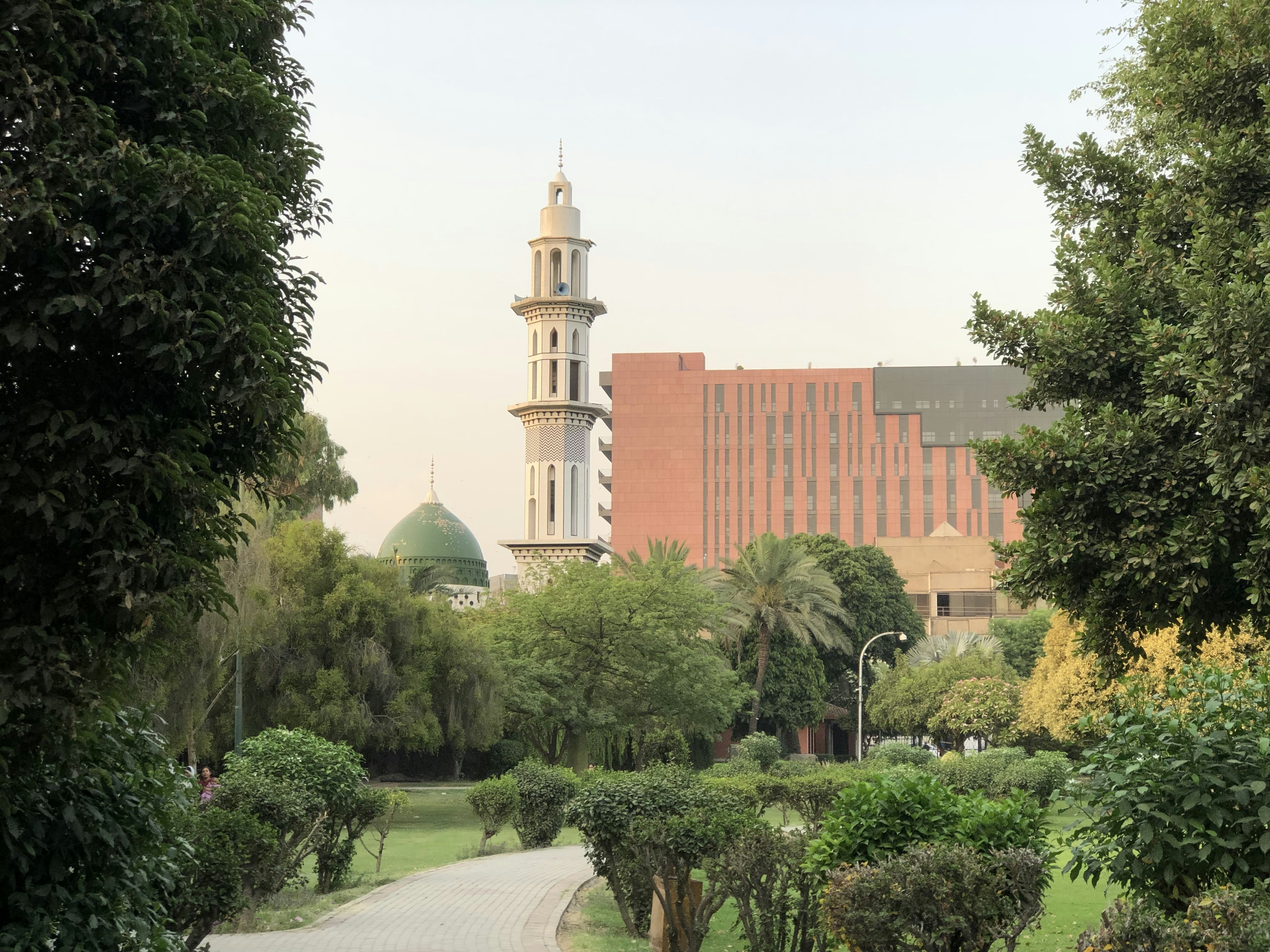 A serene park pathway leading to a striking minaret and modern building, framed by lush greenery.