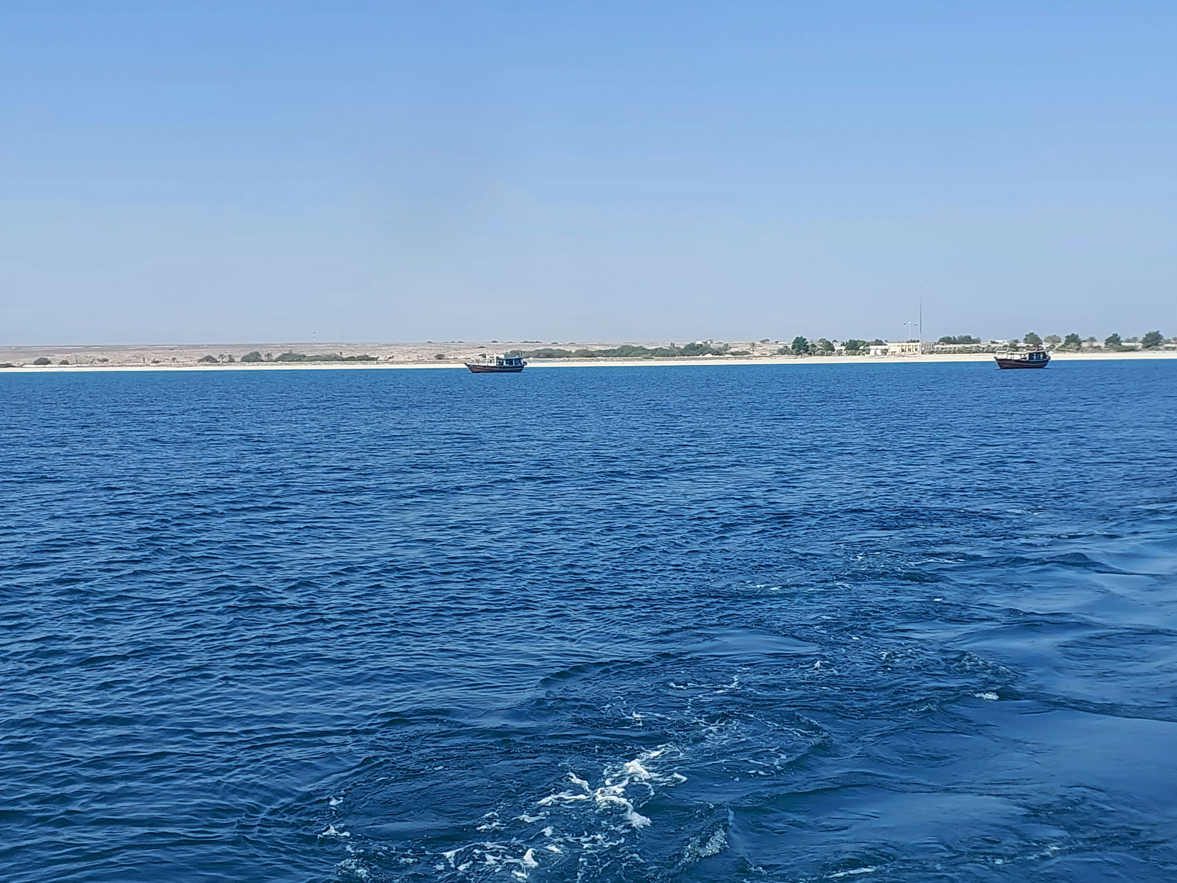 Two fishing boats gently bobbing on a calm sea, with a sandy shoreline and sparse vegetation in the background. The clear blue sky reflects the serene atmosphere.