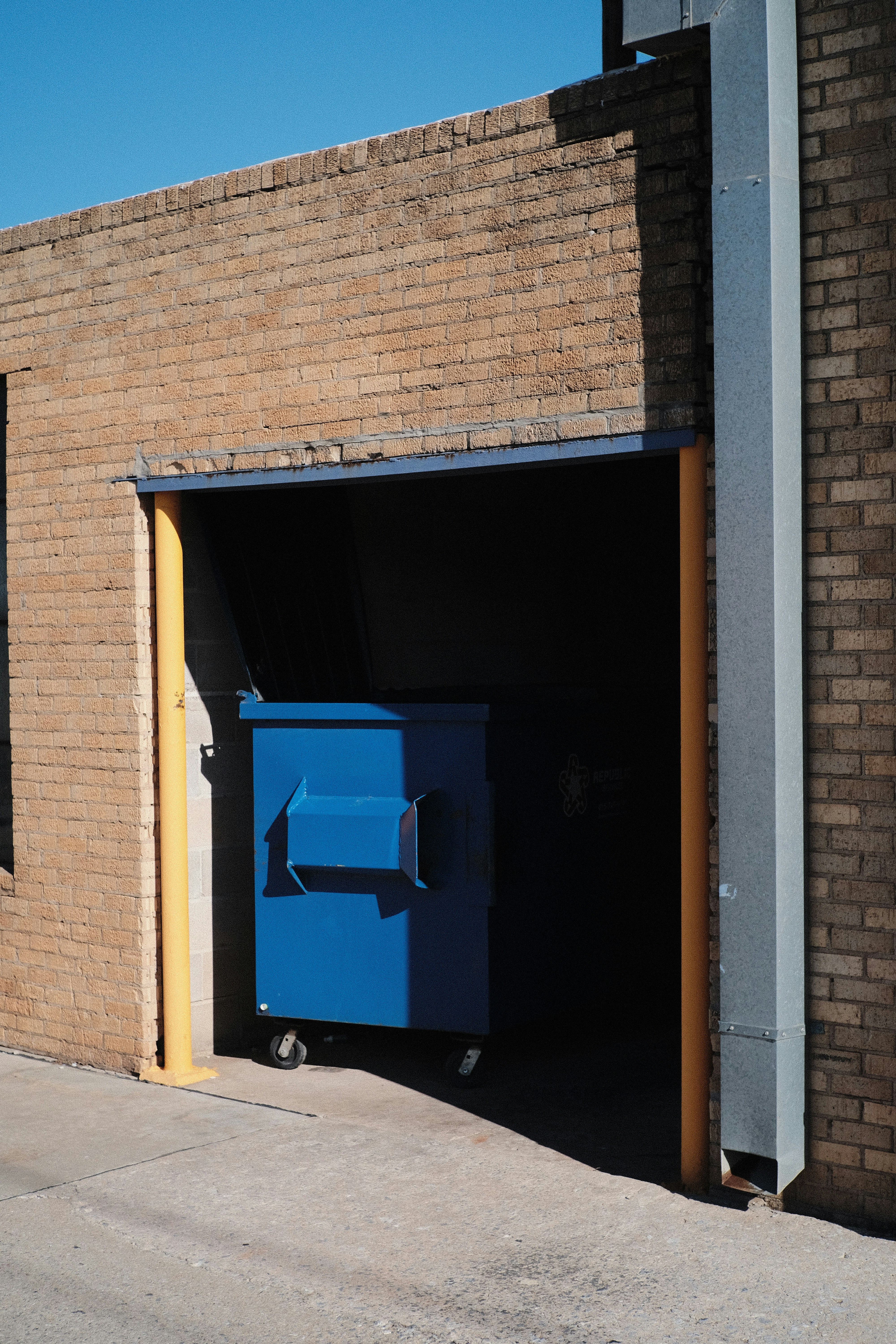 A blue dumpster partially concealed in a brick alcove, highlighting the contrast between urban decay and vibrant colors.