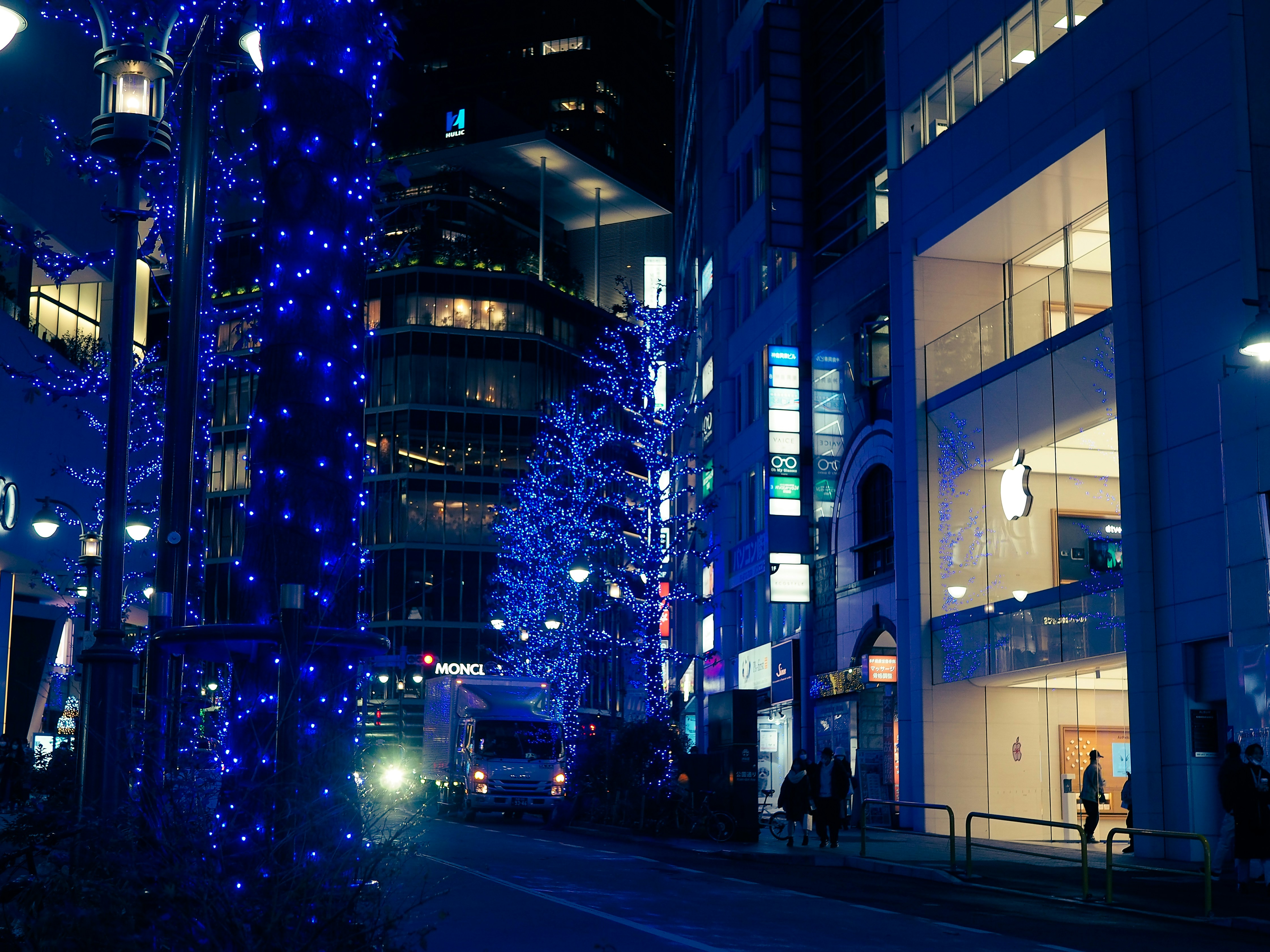 Vibrant blue lights adorn city trees near an Apple Store, creating a lively nighttime atmosphere in an urban setting.