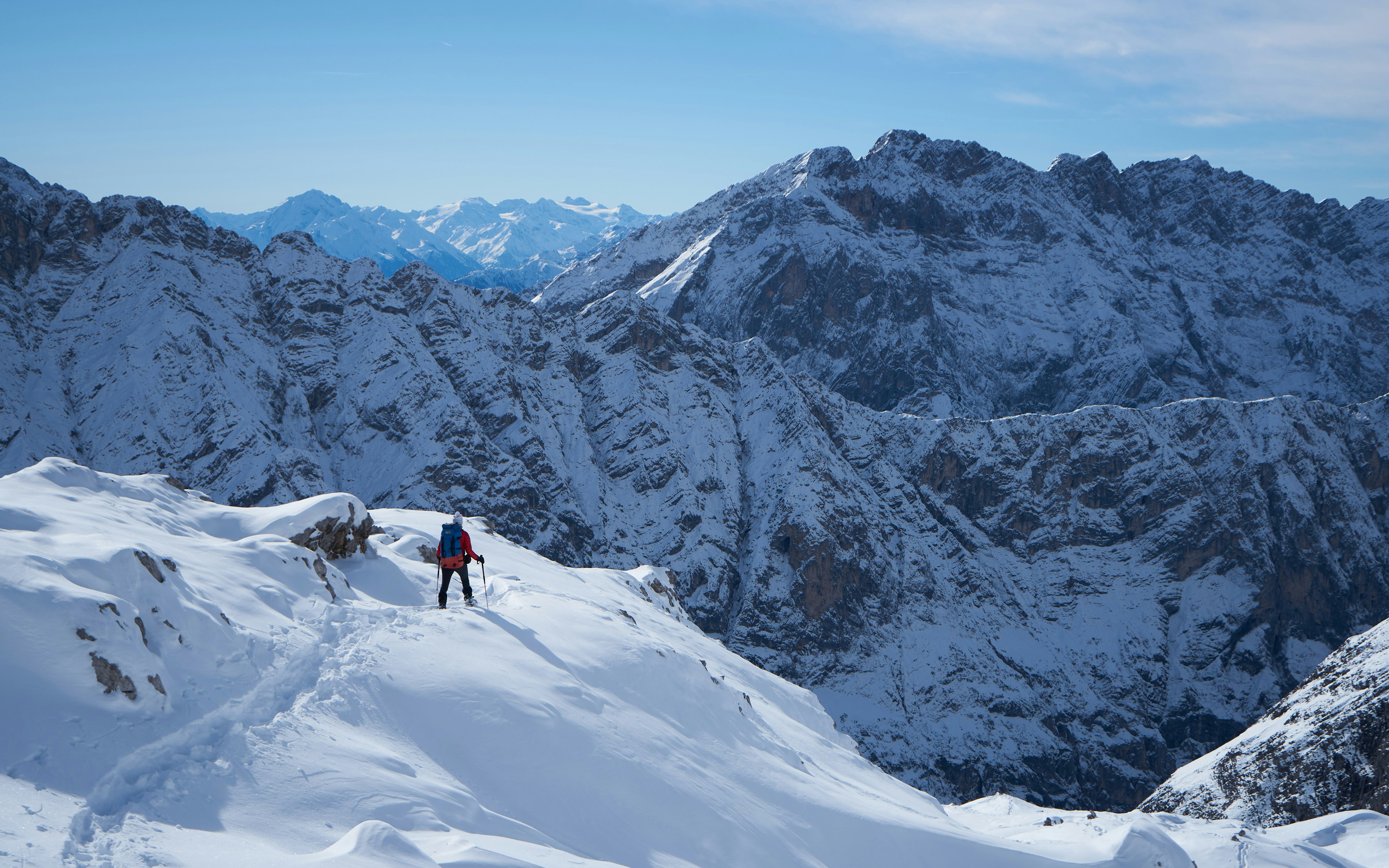 A lone hiker stands atop a snow-covered peak, surrounded by majestic mountain ranges under a clear blue sky.