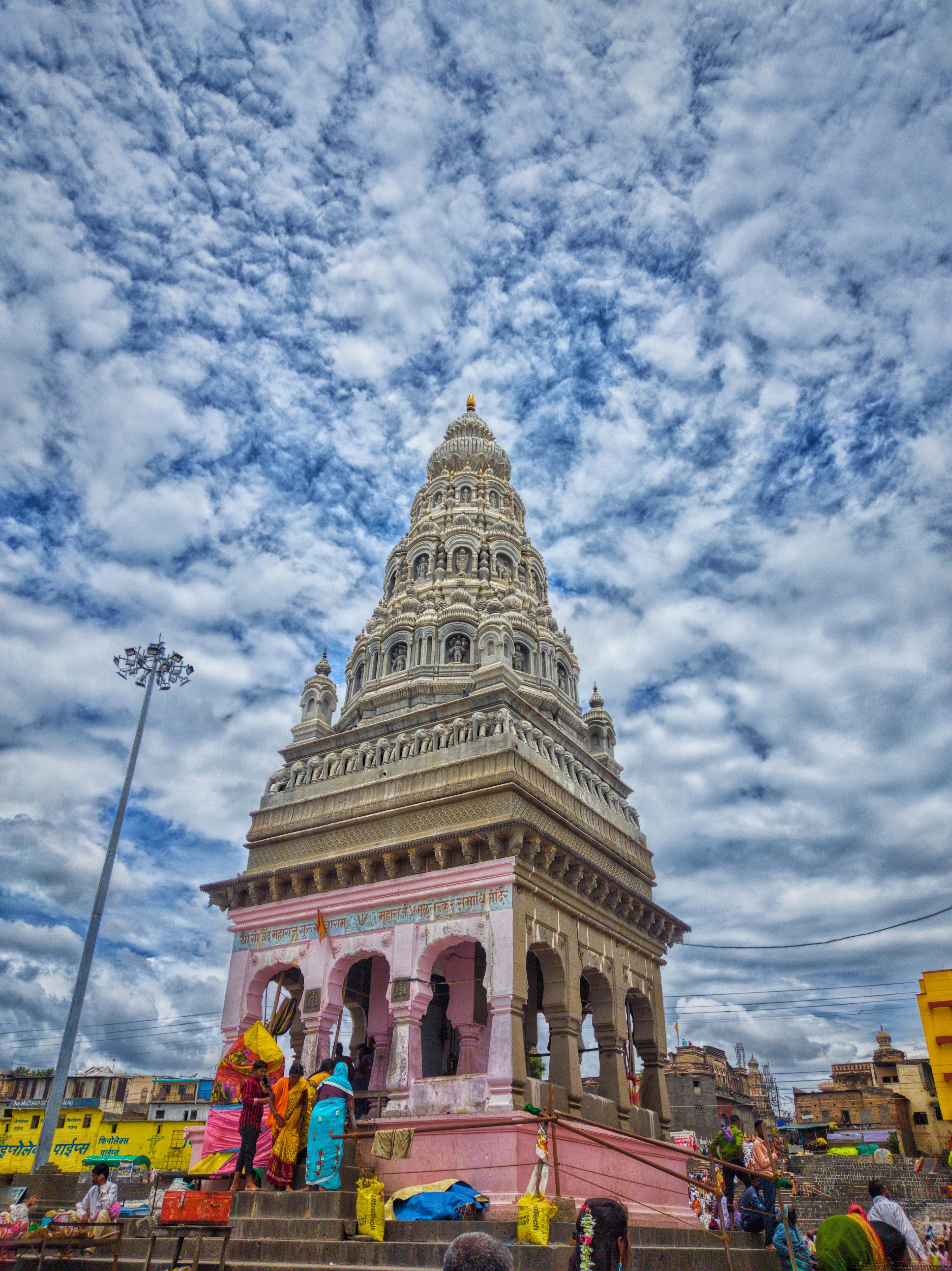 A grand temple adorned with intricate architecture stands under a dynamic sky, bustling with devotees and vibrant colors. The scene captures the essence of cultural devotion and architectural beauty.