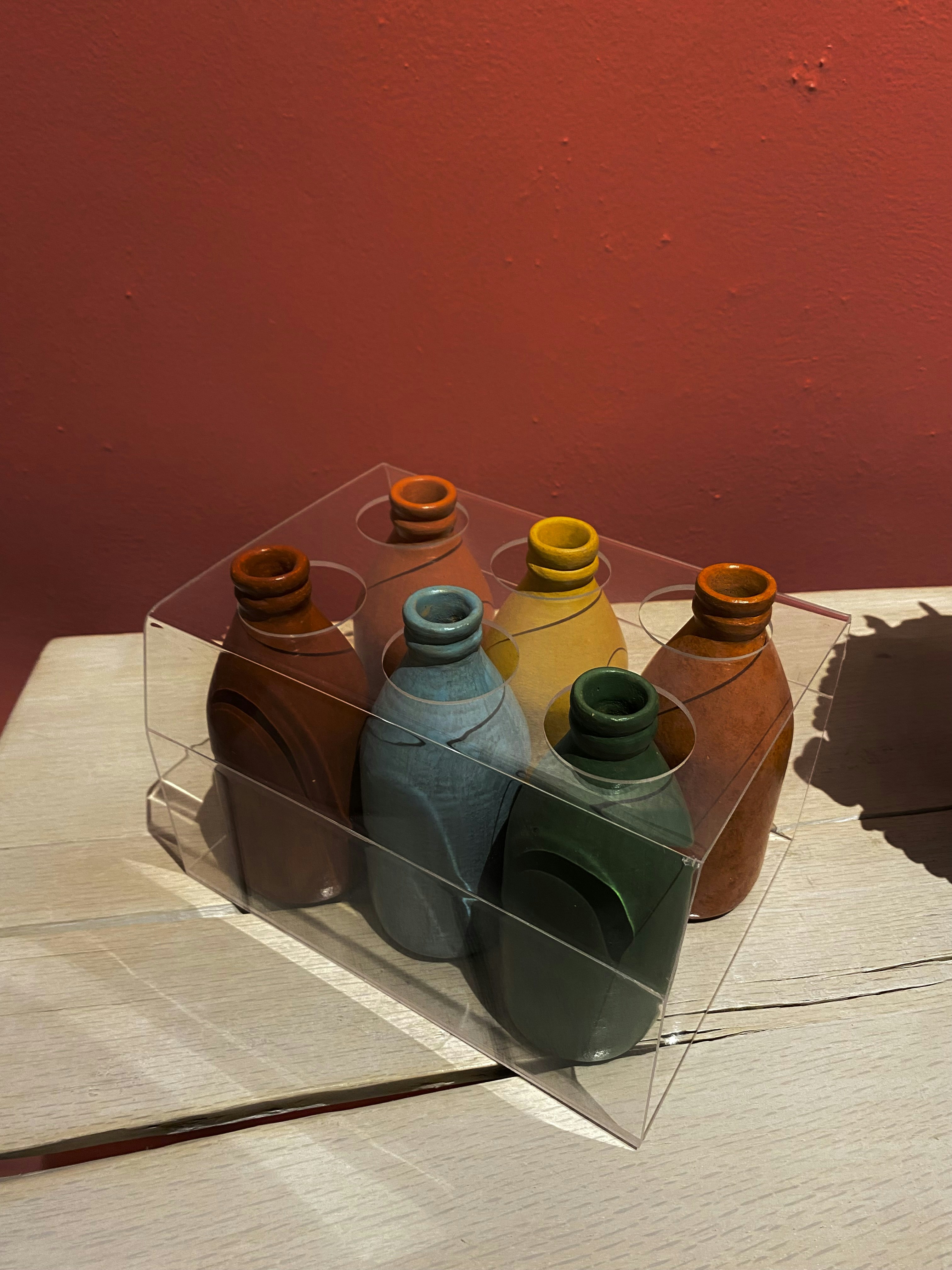 Six Colored Ceramic Bottles Nestled in a Transparent Carrier on a Sunlit Wood Surface