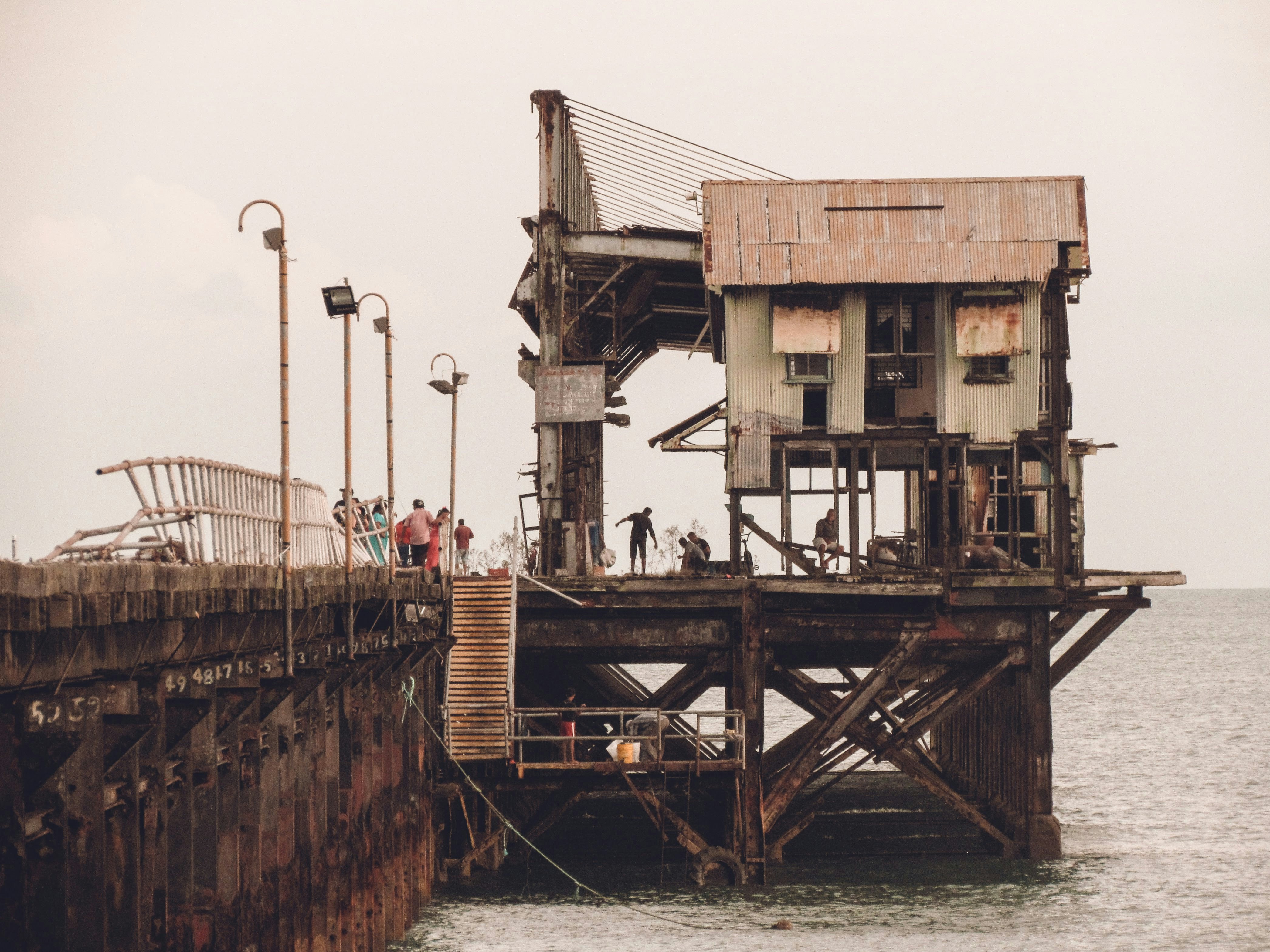 An old pier structure with a rustic building at the end, surrounded by calm waters. People gather, adding life to the historical scene.