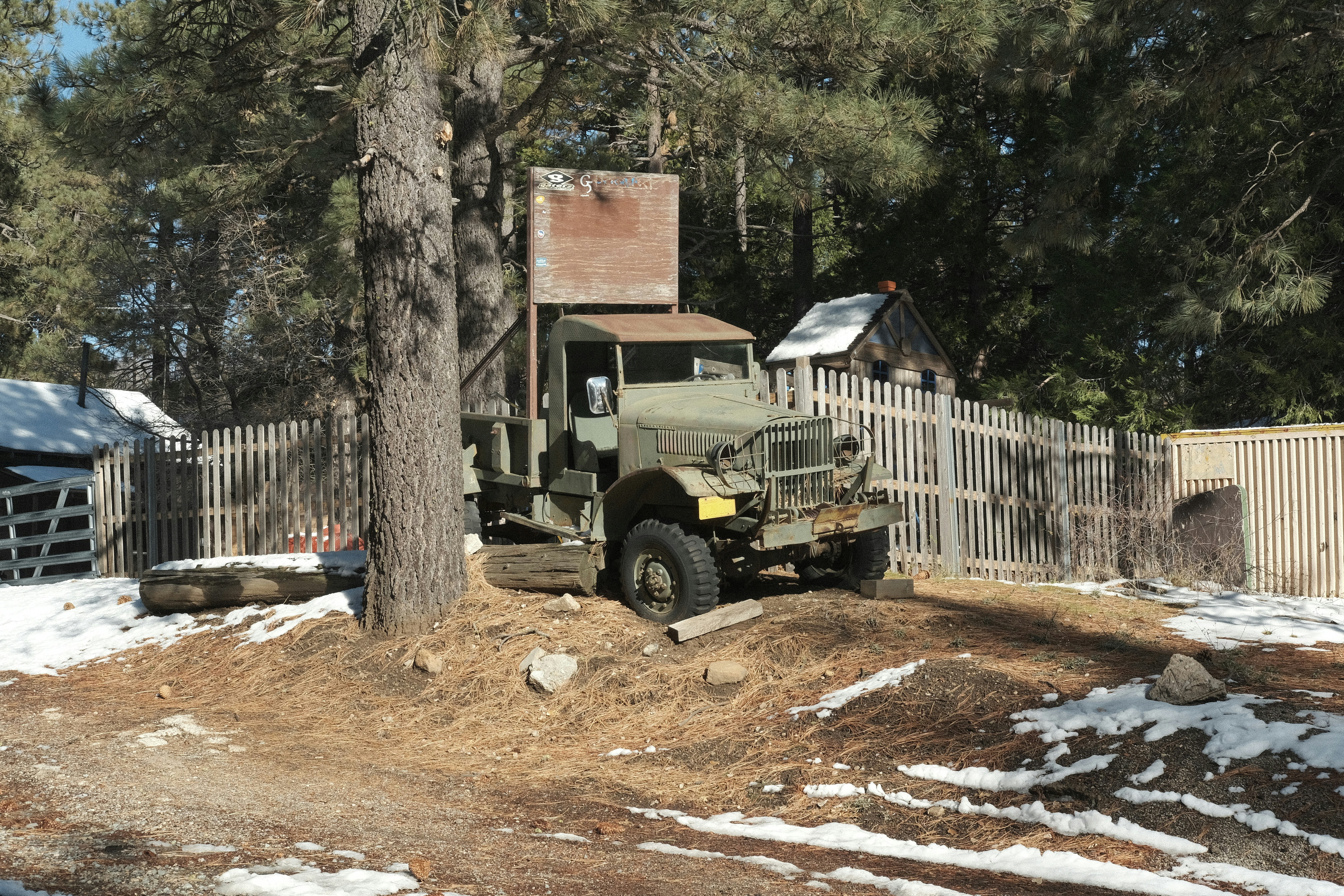 An old military truck sits prominently on a snowy patch of ground, surrounded by trees and rustic fencing, showcasing its weathered charm.