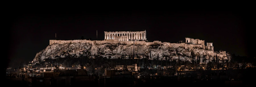 The Parthenon sits brightly lit atop the Acropolis against a deep black night sky, with the rocky hillside and city lights below.