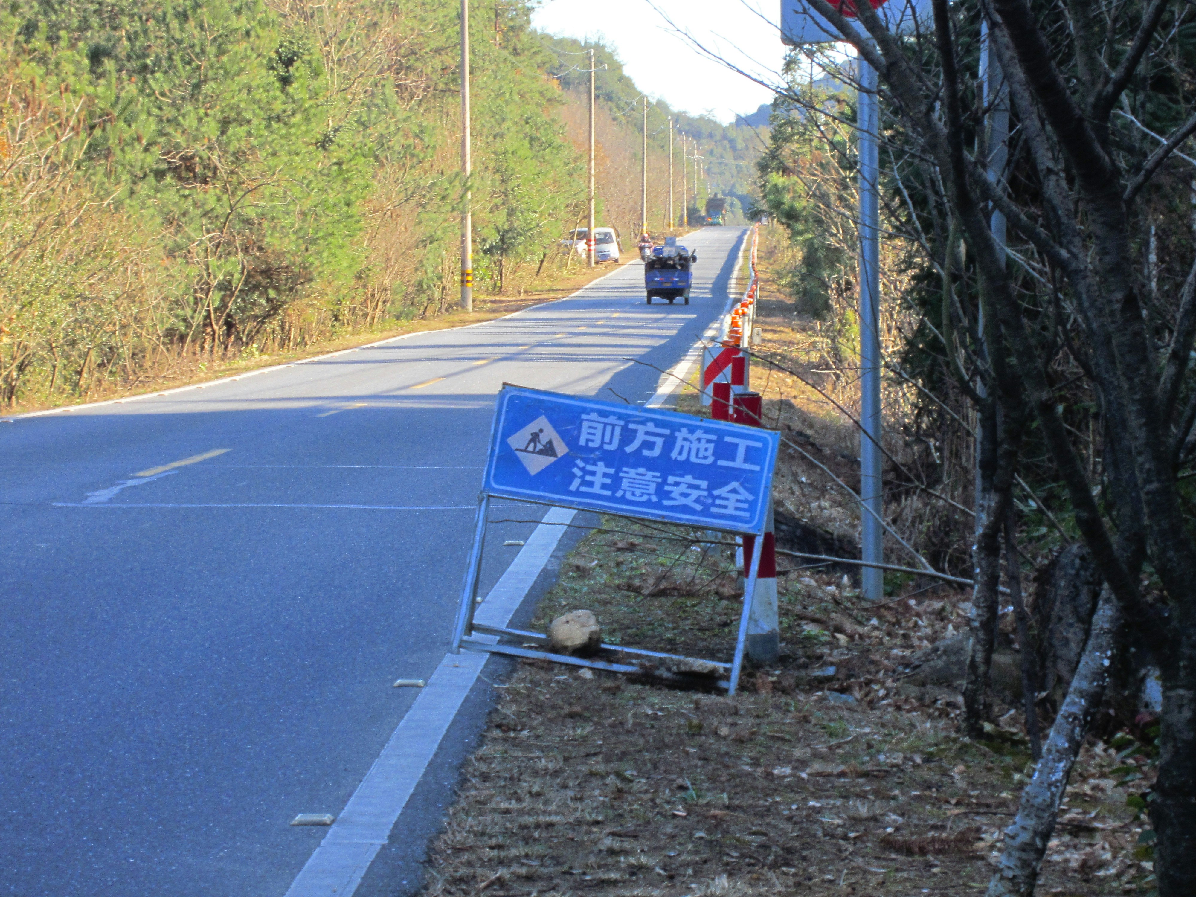 A construction warning sign leaning by the roadside, surrounded by trees and a clear road ahead.