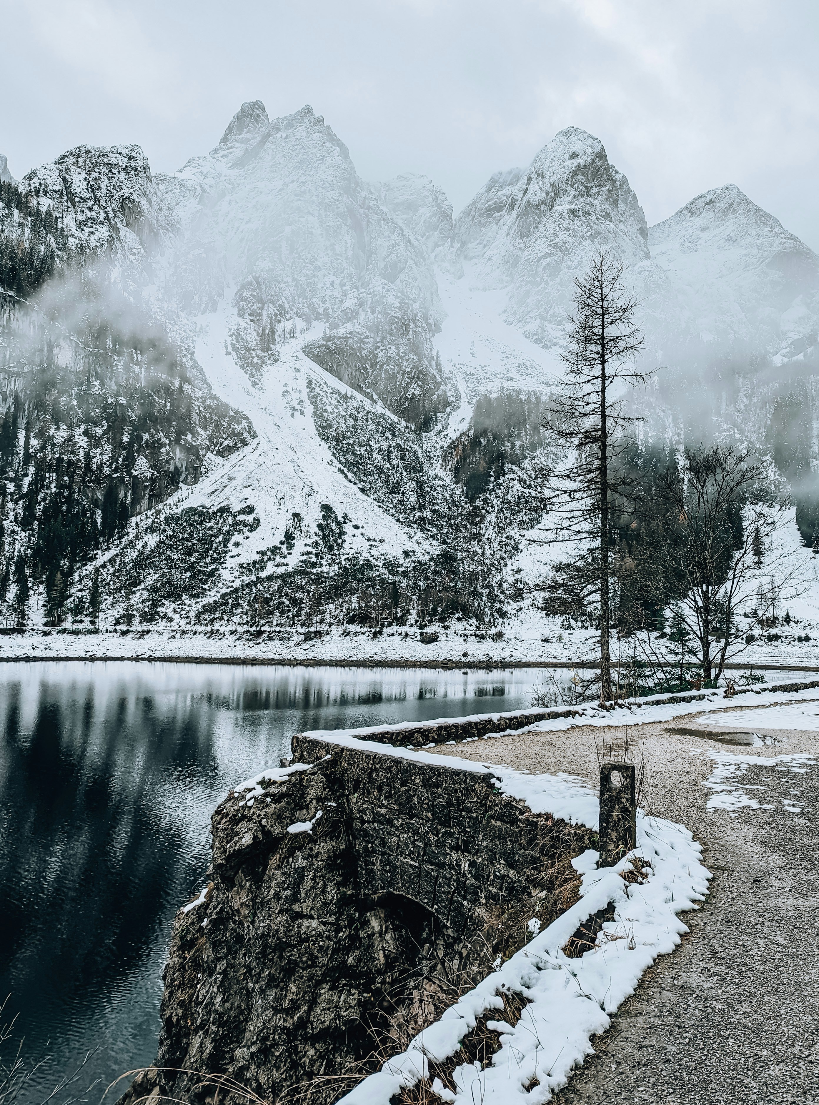 Snow-covered mountains loom over a tranquil lake, with a winding path leading through a winter landscape.