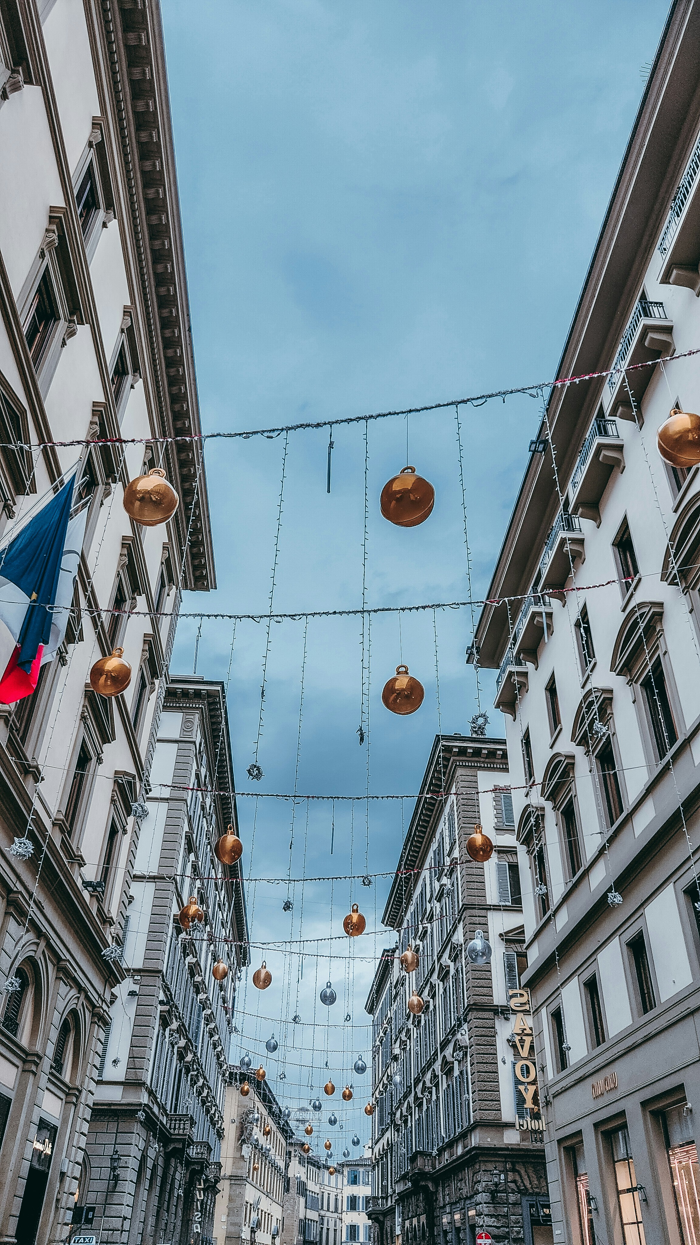 Festive decorations hang between historic buildings in a city street, under a cloudy sky. The scene captures the essence of urban celebrations.