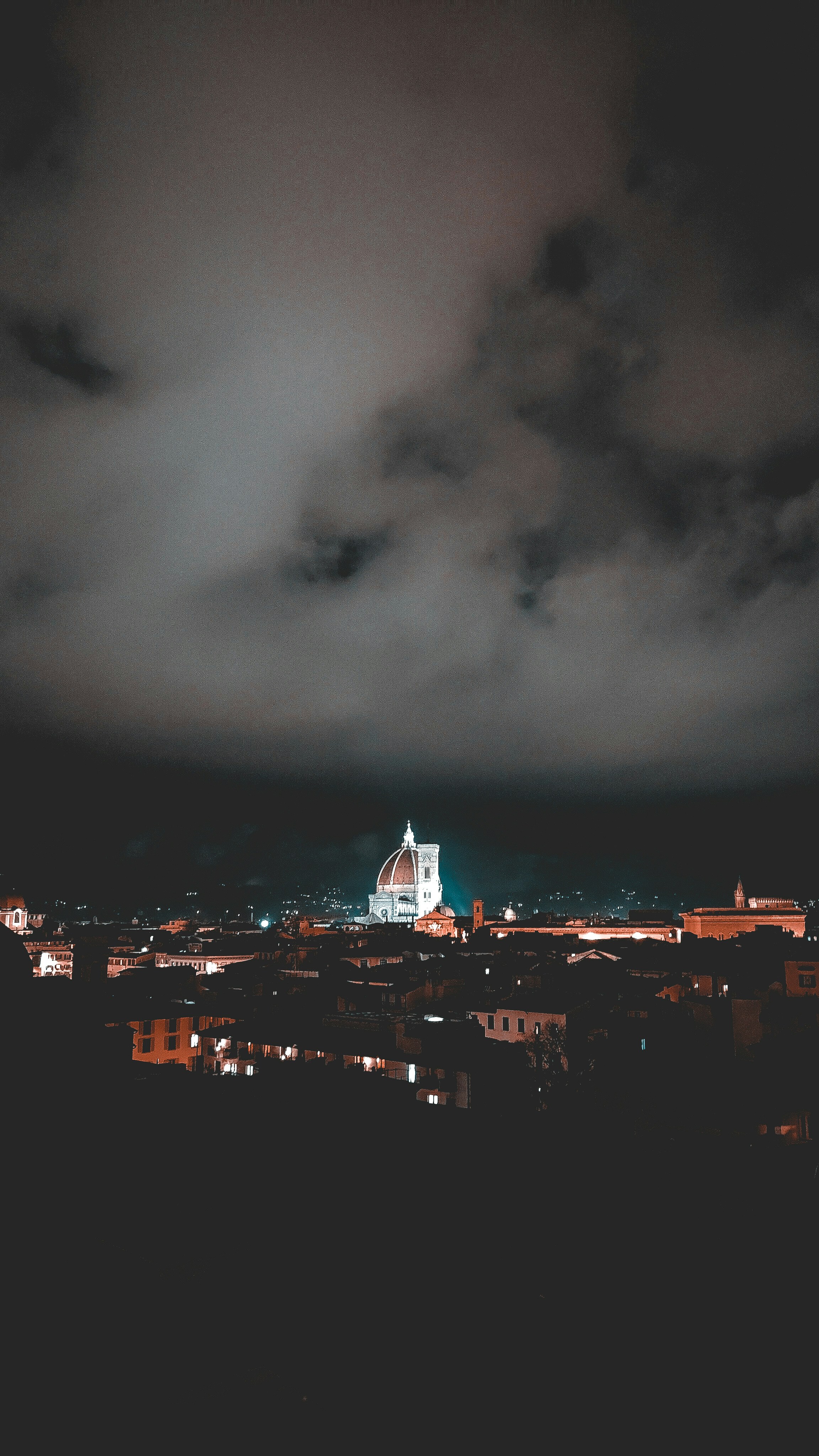 Night cityscape with a brightly lit cathedral dome rising above rooftops beneath a cloud-filled sky.