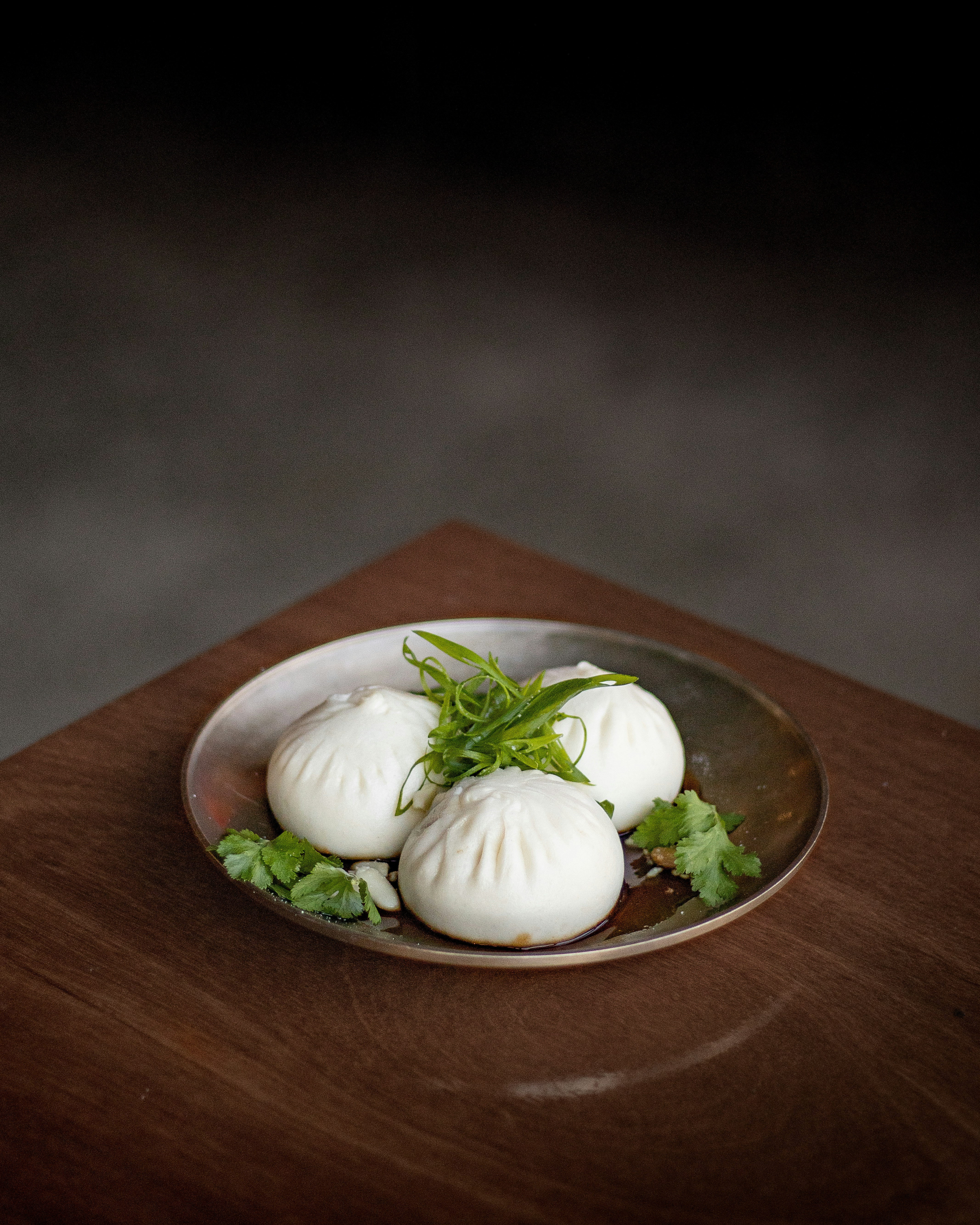Three fluffy steamed buns garnished with green onions and cilantro on a round plate, set against a dark background.