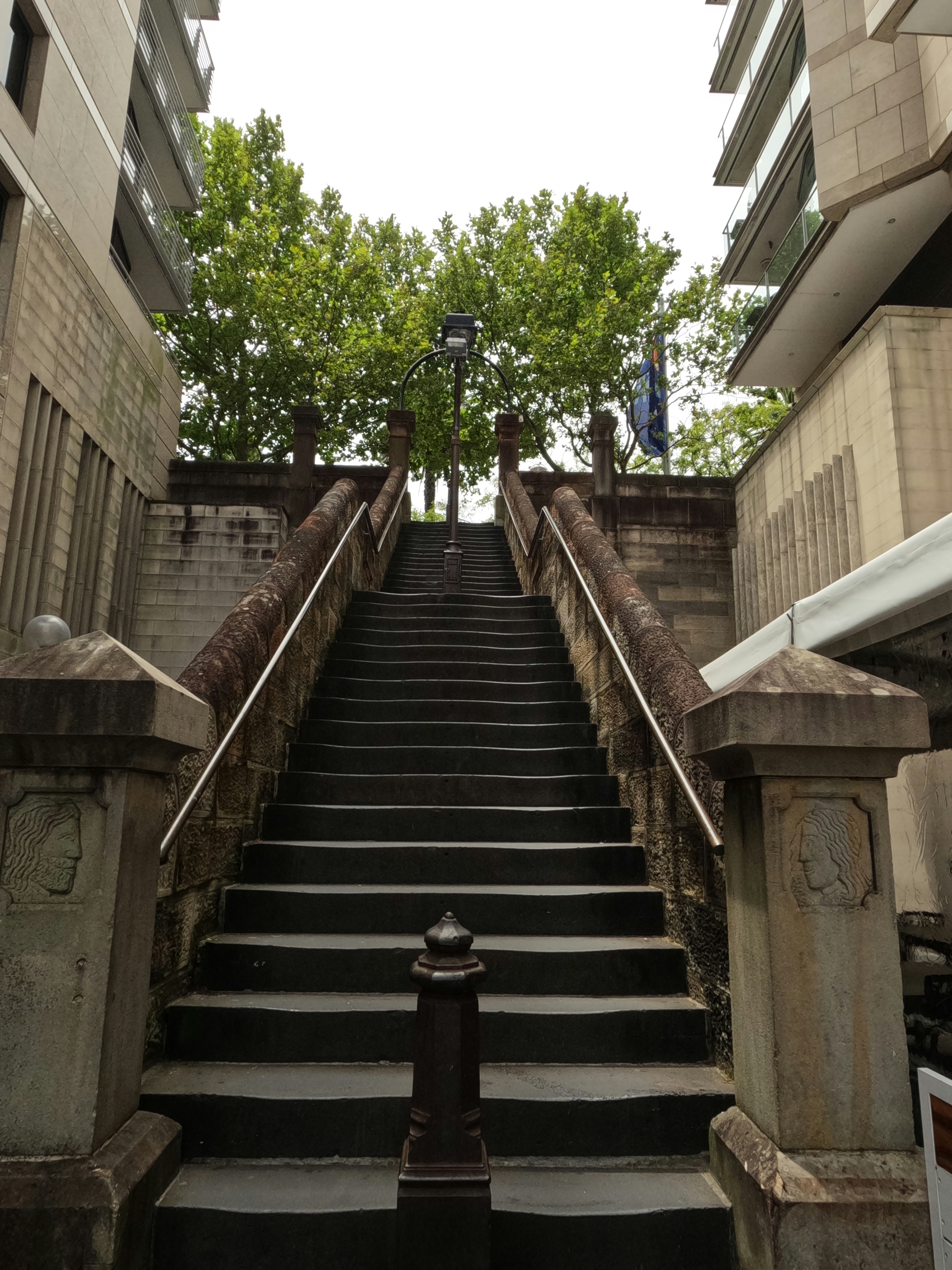 Stone staircase flanked by weathered balustrades climbs toward a tree-canopied plaza in an urban setting. A lone lamp post anchors the central axis as daylight softens the stone textures.