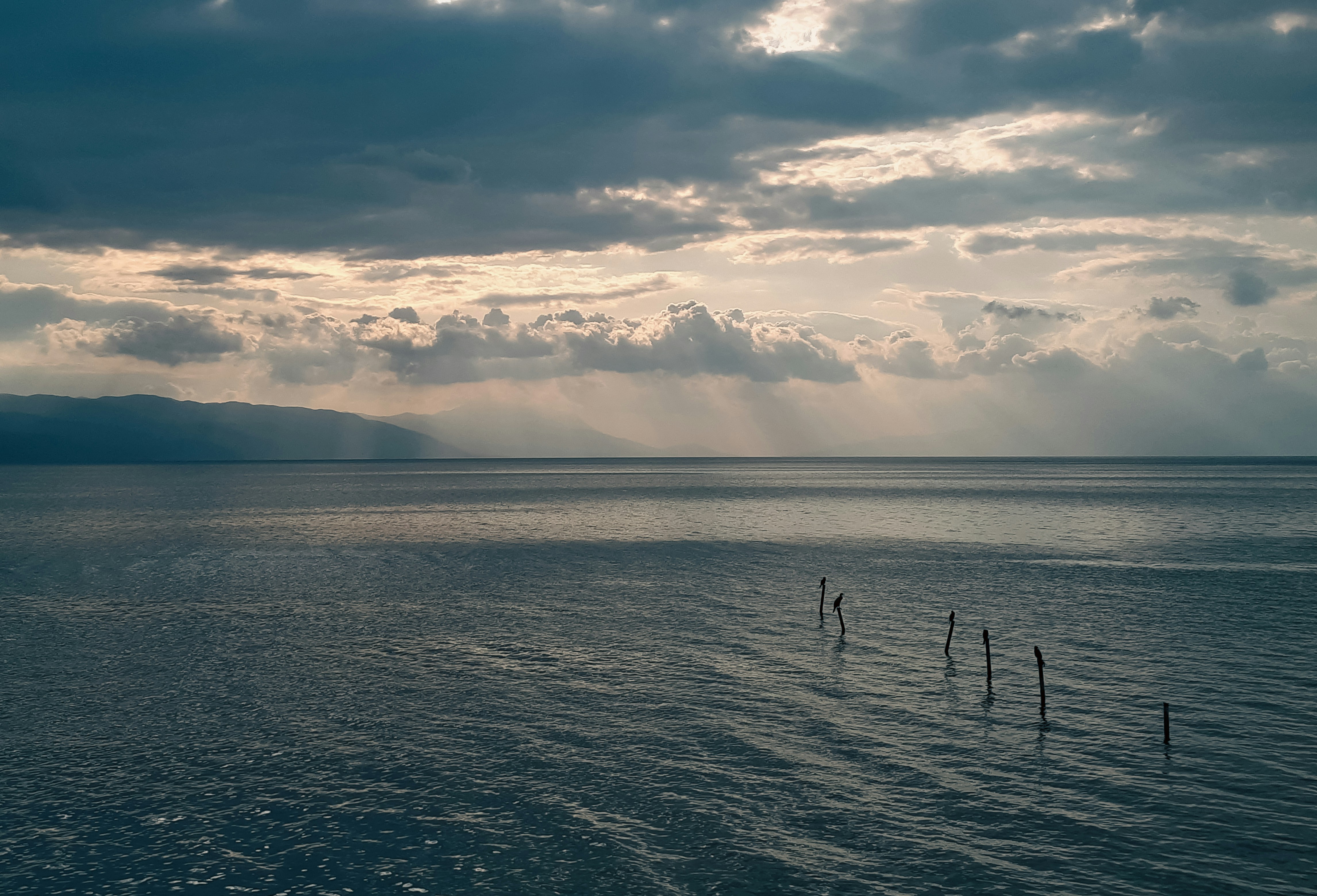 Serene seascape featuring gentle waves and distant mountains under a dramatic sky with rays of light breaking through clouds.