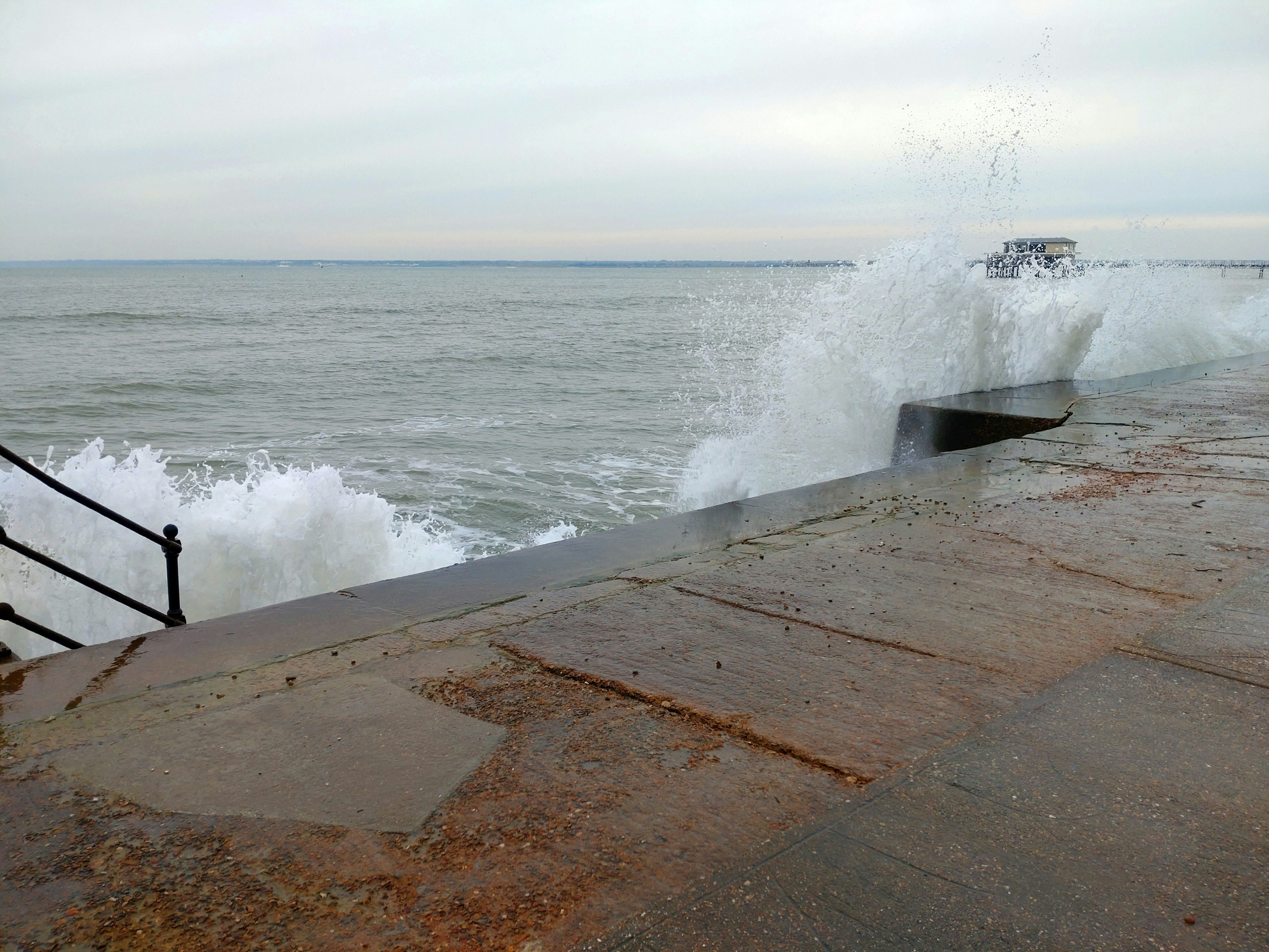 Choppy sea along a rust-stained seawall with a large spray surge breaking near the right, foregrounds a textured concrete ledge and railing. A distant horizon anchors the composition.