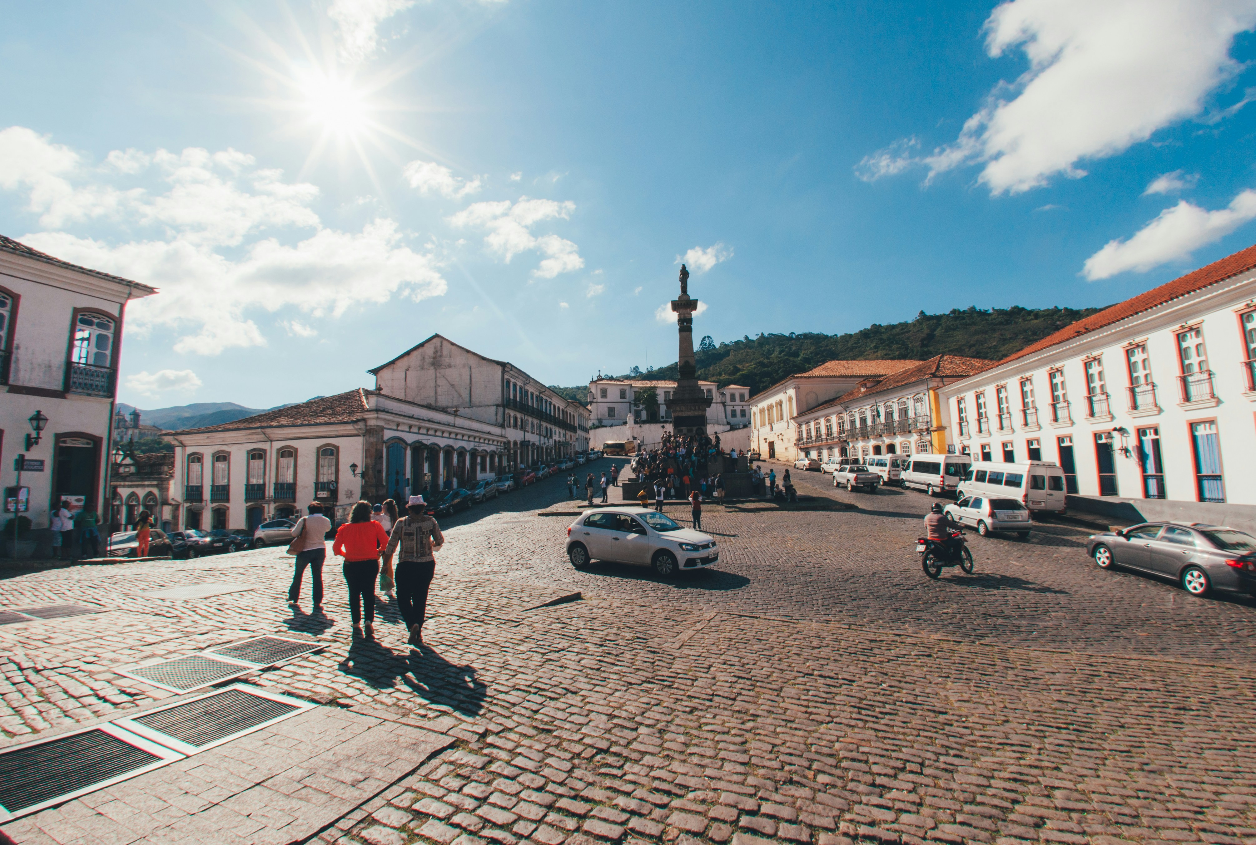 People walking along a cobblestone street in a sunlit town square with historic architecture.
