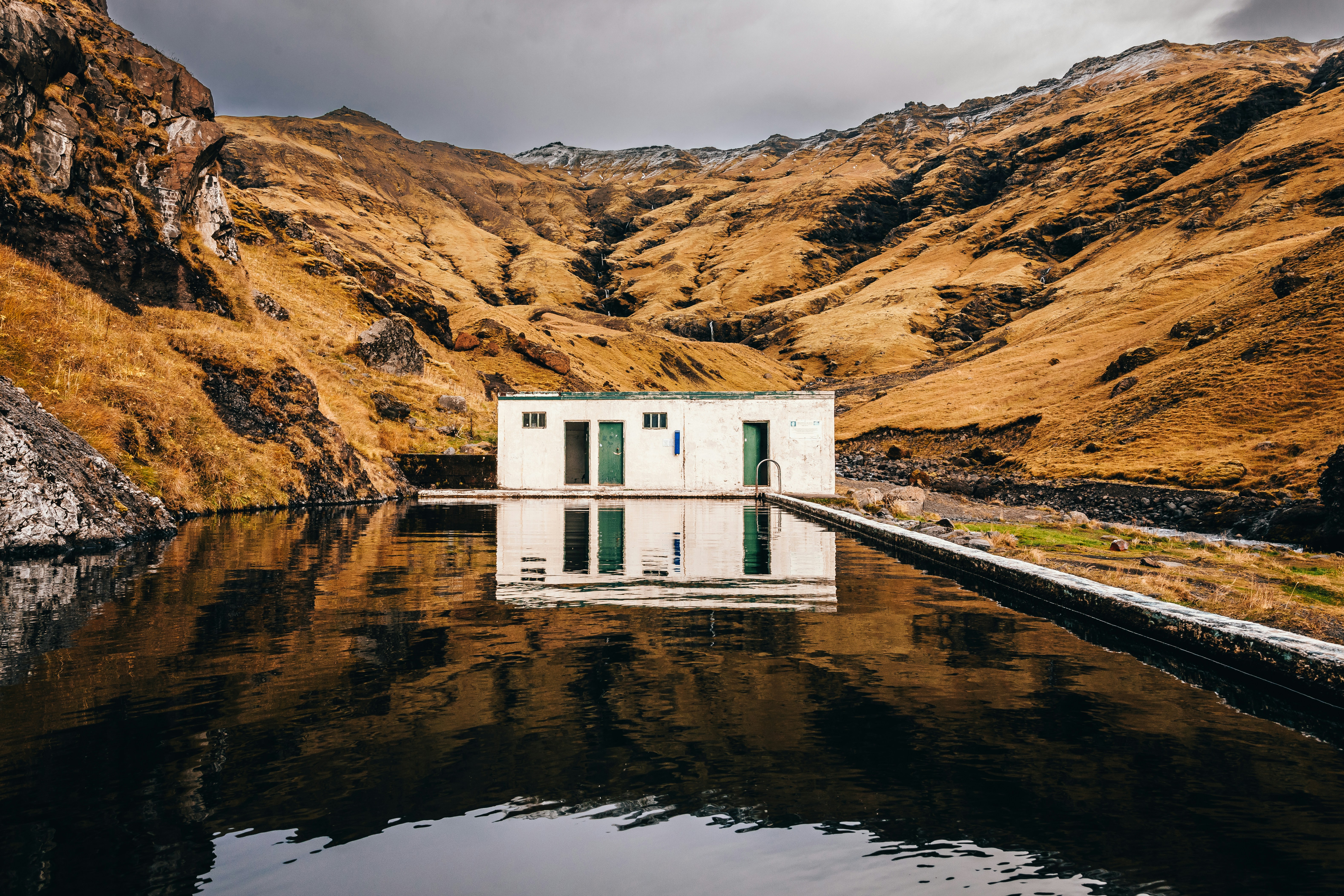 An abandoned building reflects in a still body of water, surrounded by golden hills under a moody sky.