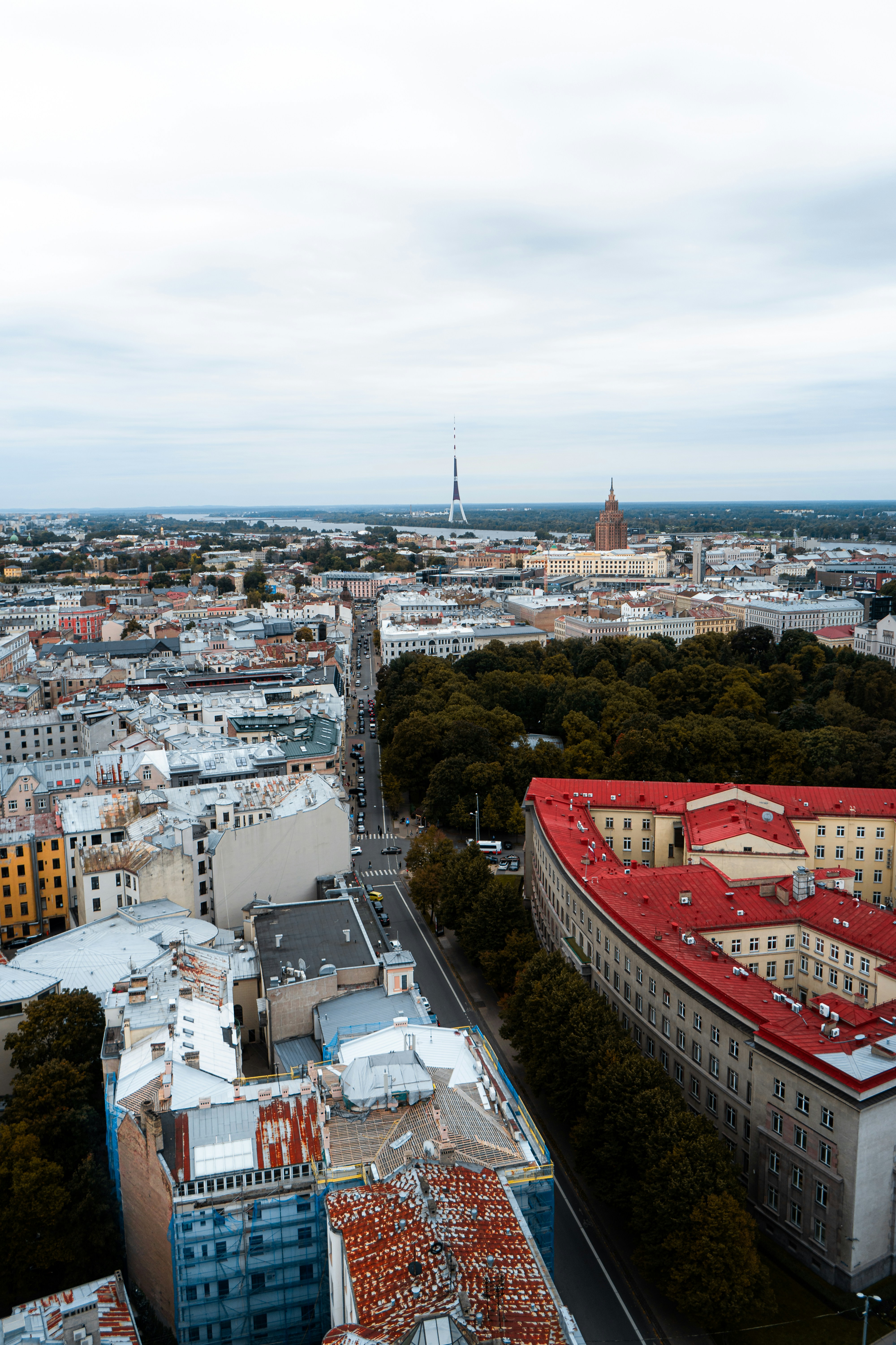 Aerial view of a city with a blend of historical and modern architecture, showcasing rooftops and urban greenery. The skyline features a prominent tower and church spire.